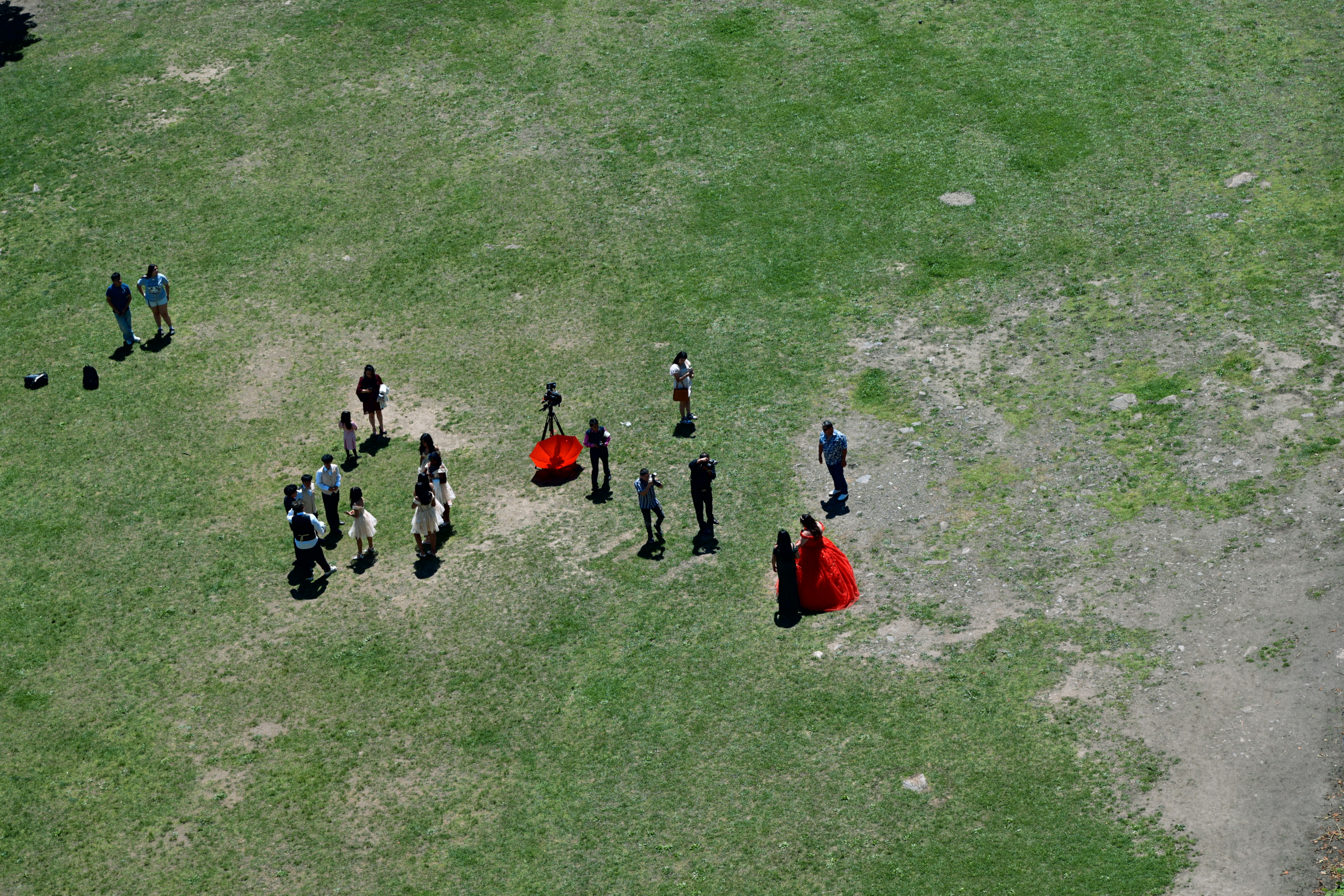 A drone view shows a cluster of people gathered on a grassy, patchy field with two red objects adding color to the scene.