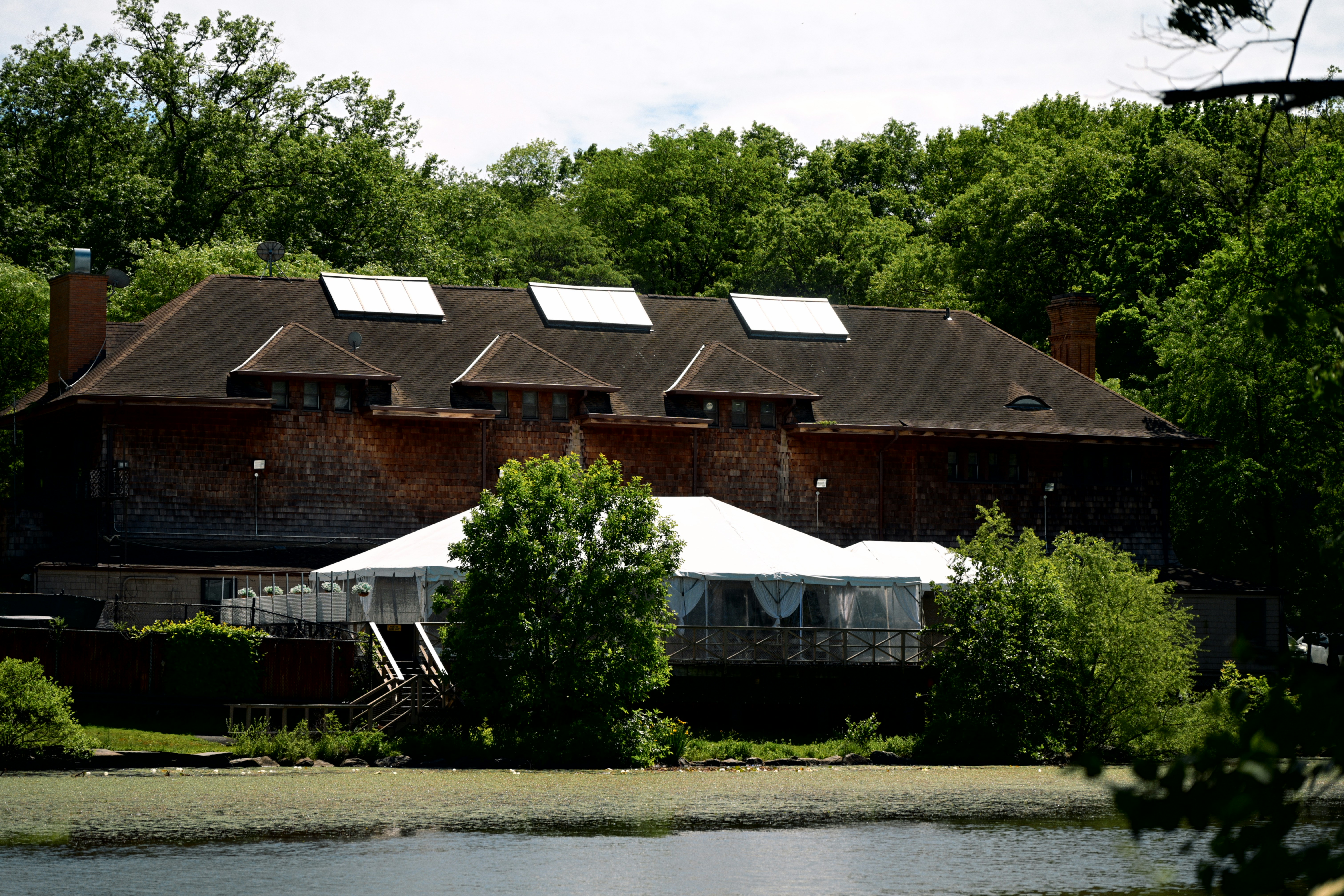 Large building and white tent beside water, surrounded by trees.