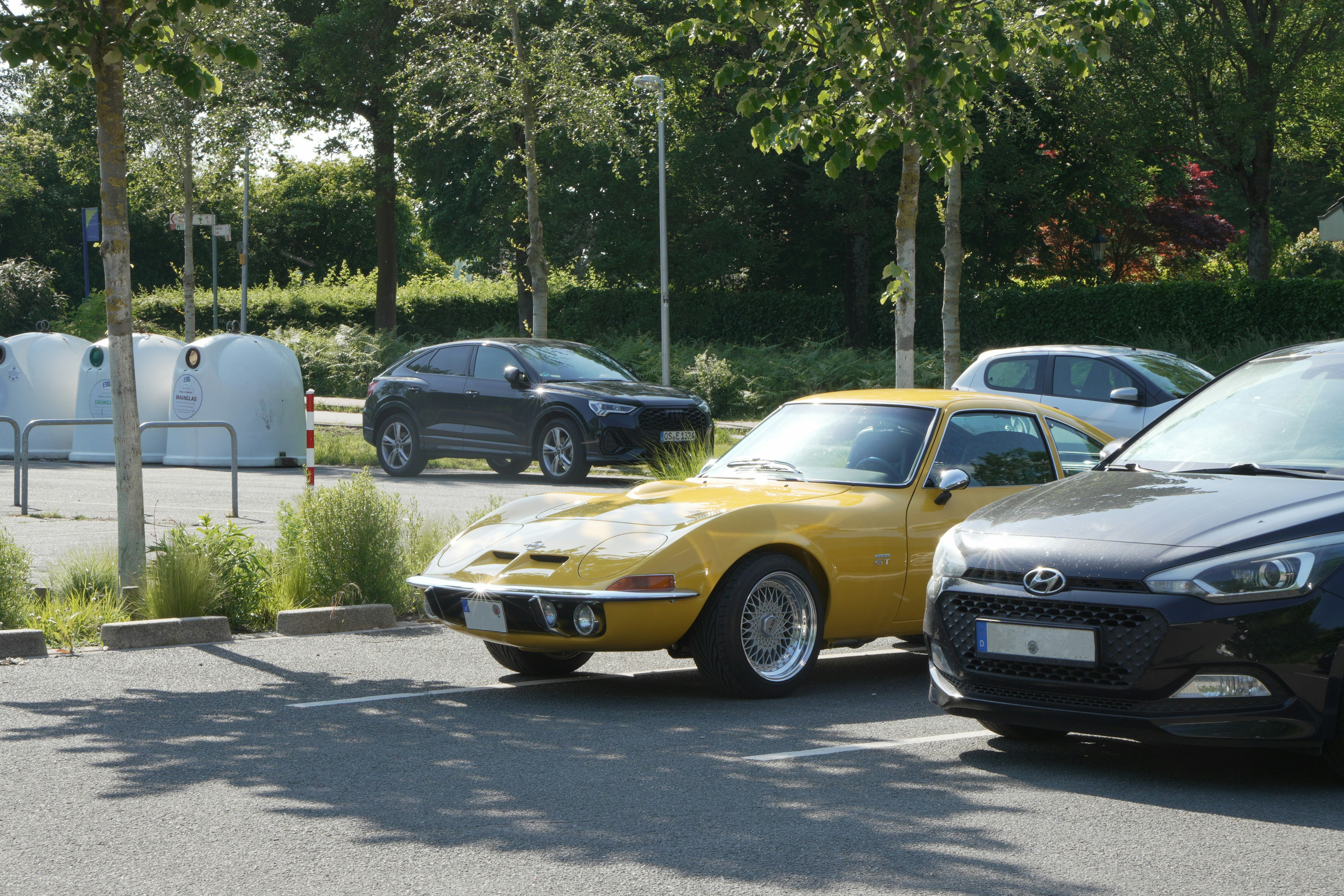 Two classic cars parked in a sunlit lot with trees and modern vehicles nearby, highlighting the yellow vintage coupe as the focal point.