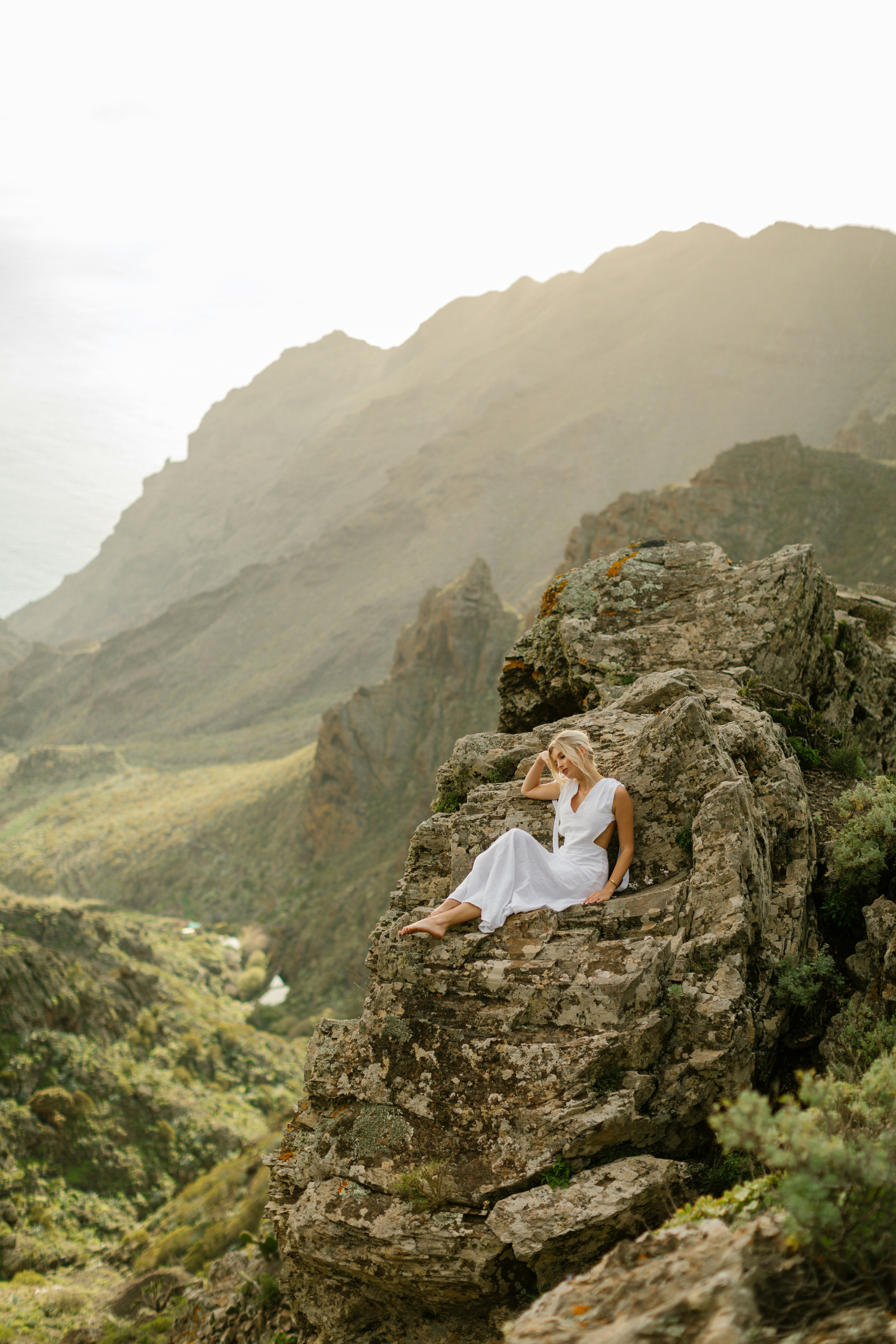 Clifftop Moment: A Woman in White Resting Amid Gnarled Rocks with Ocean Haze