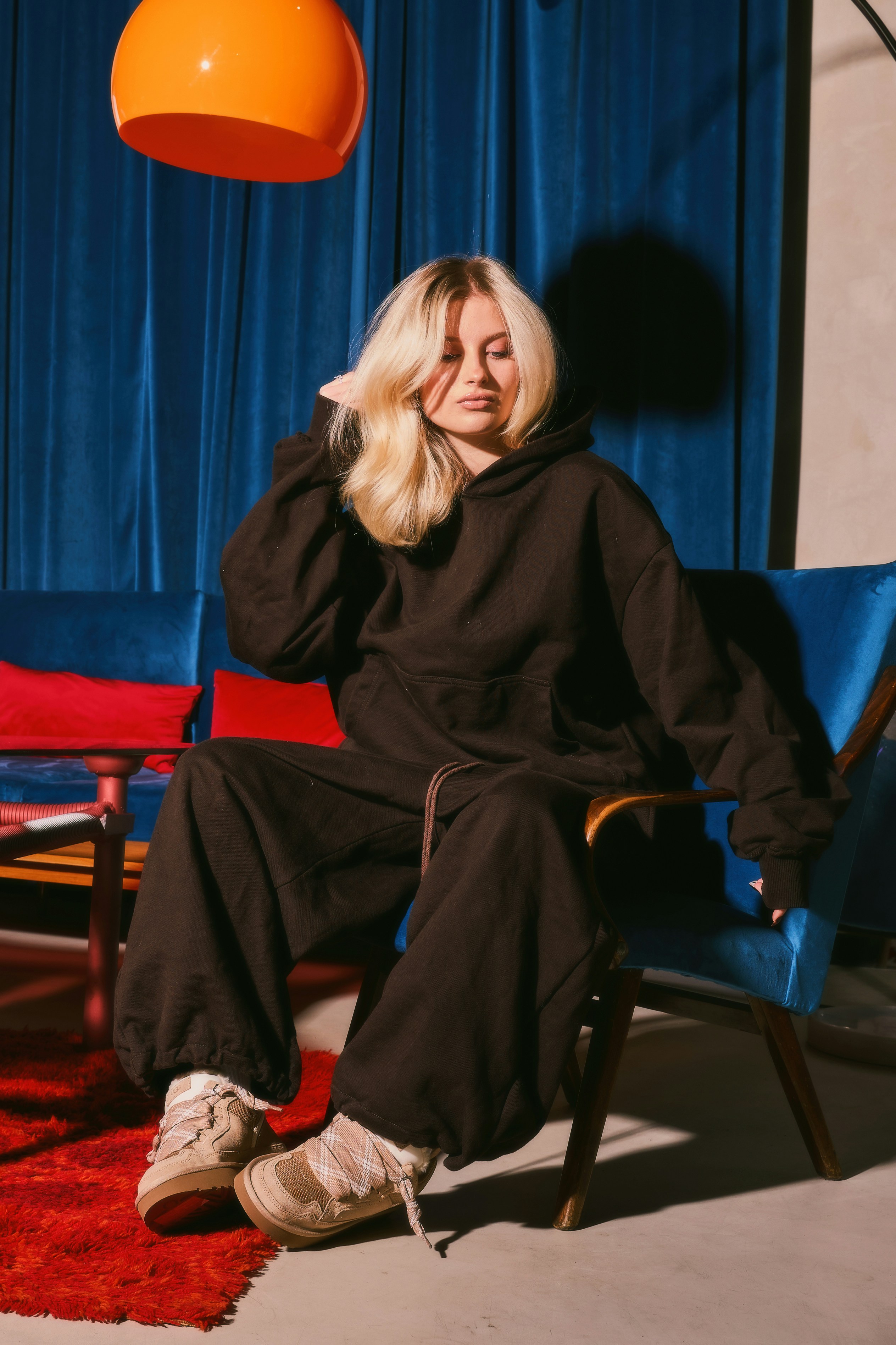 Blonde in a Black Hoodie lounging on a velvet arrangement under an orange pendant