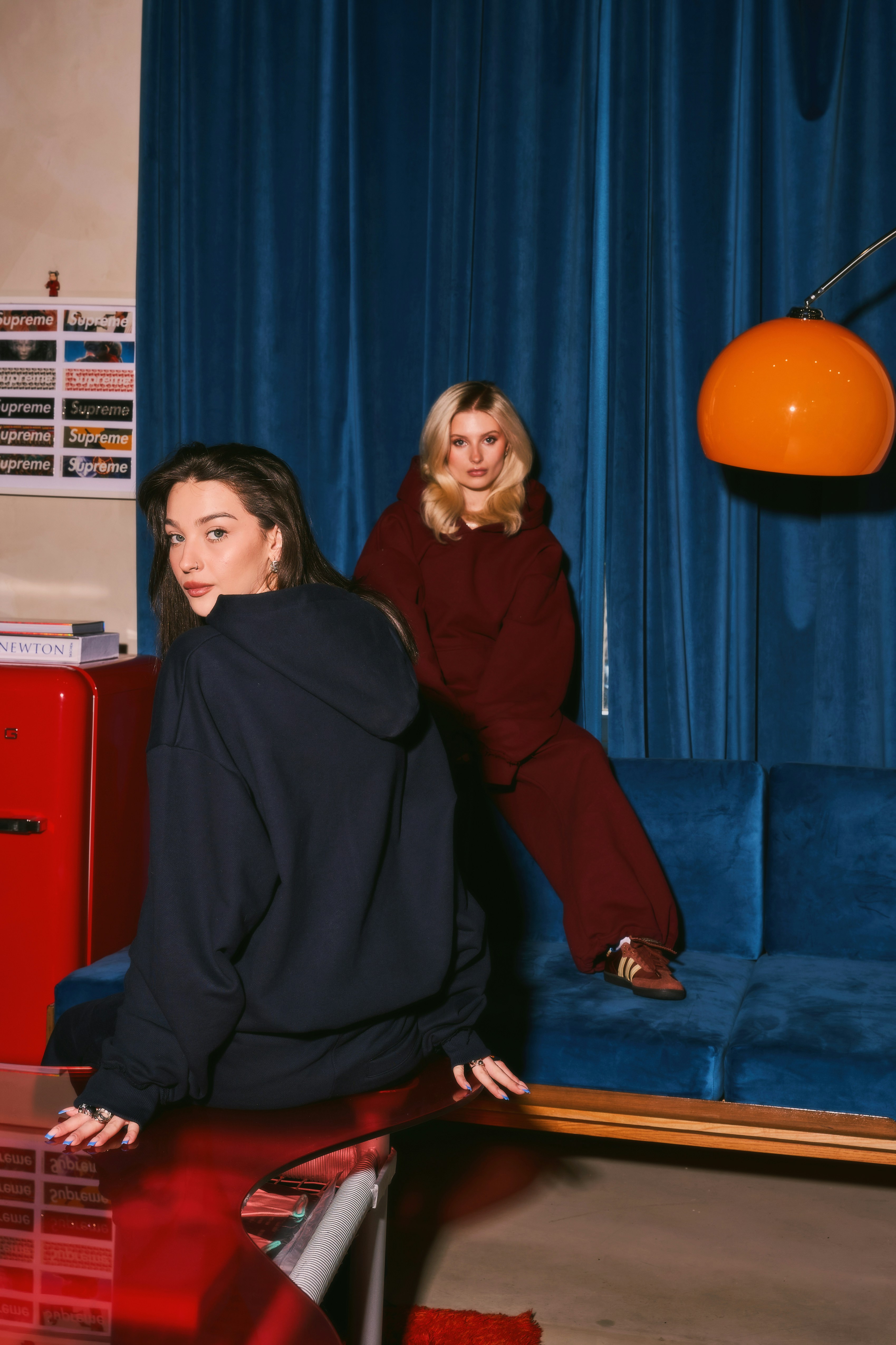 Two figures in a retro lounge with a blue velvet backdrop and a bold orange lamp