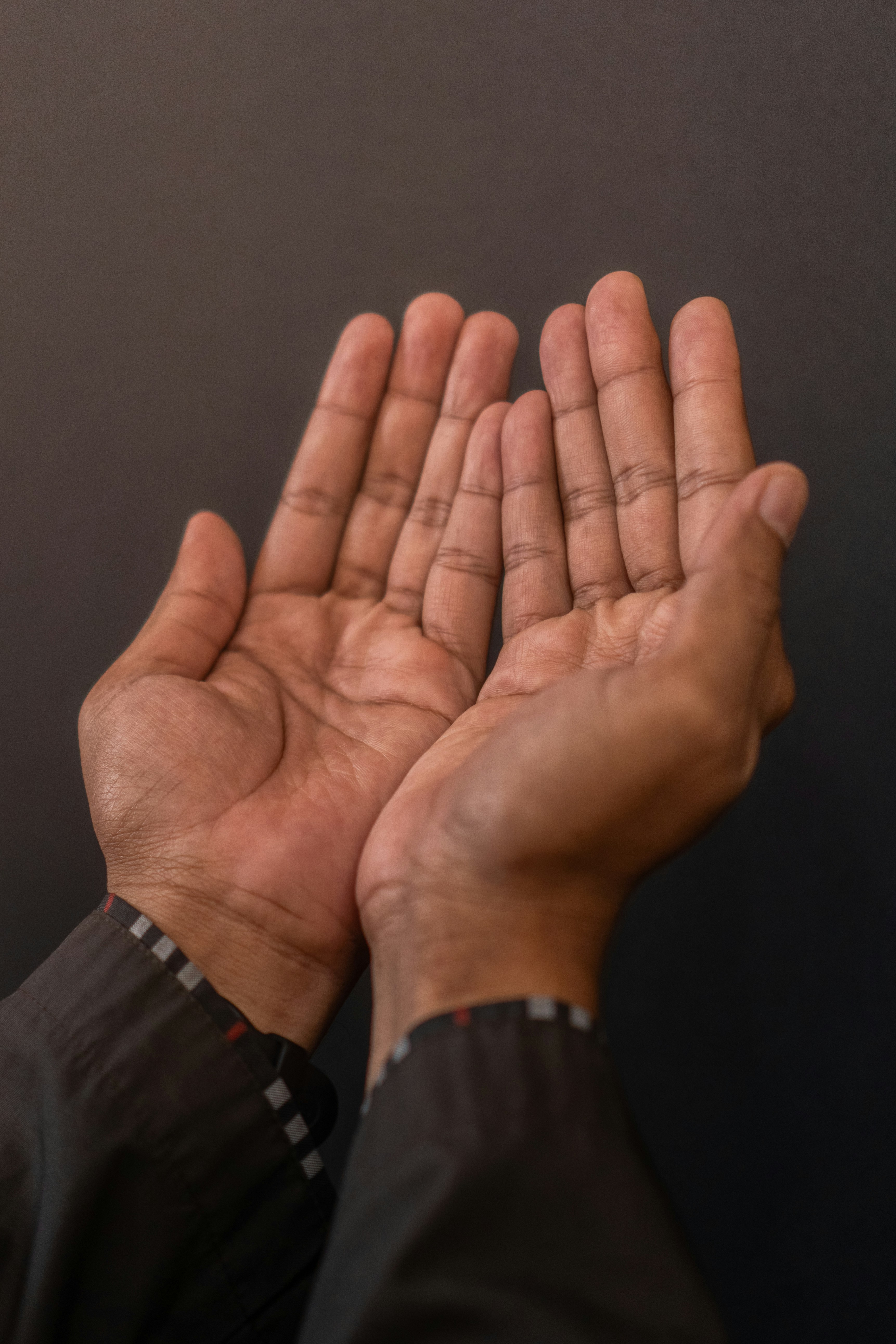Close-up of two dark-skinned hands held open against a matte black background; sleeves suggest a casual, restrained portrait or gesture-centered composition.