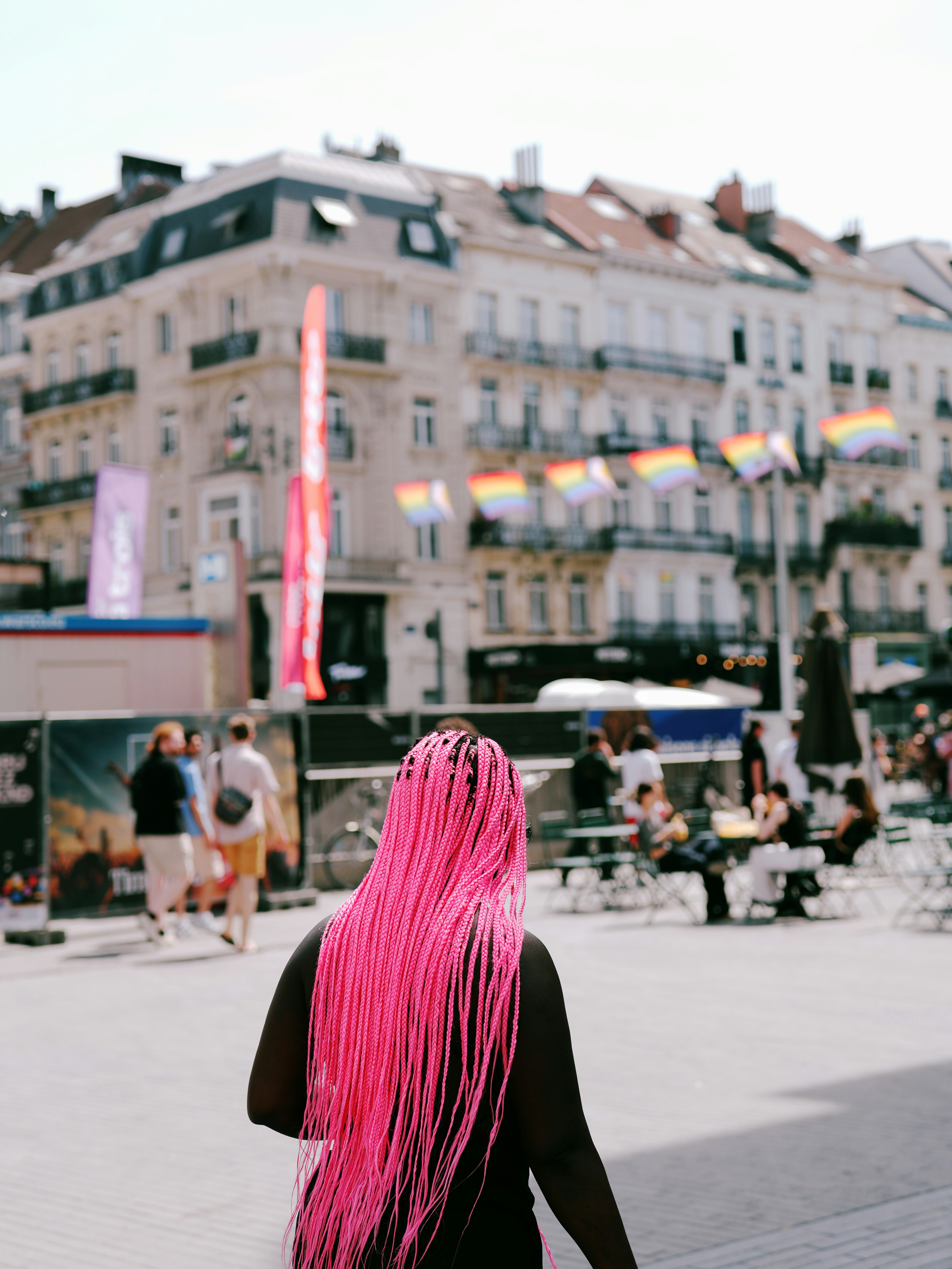 A figure with bright pink braids stands in the square as rainbow flags catch the wind across the Belle Époque façades.