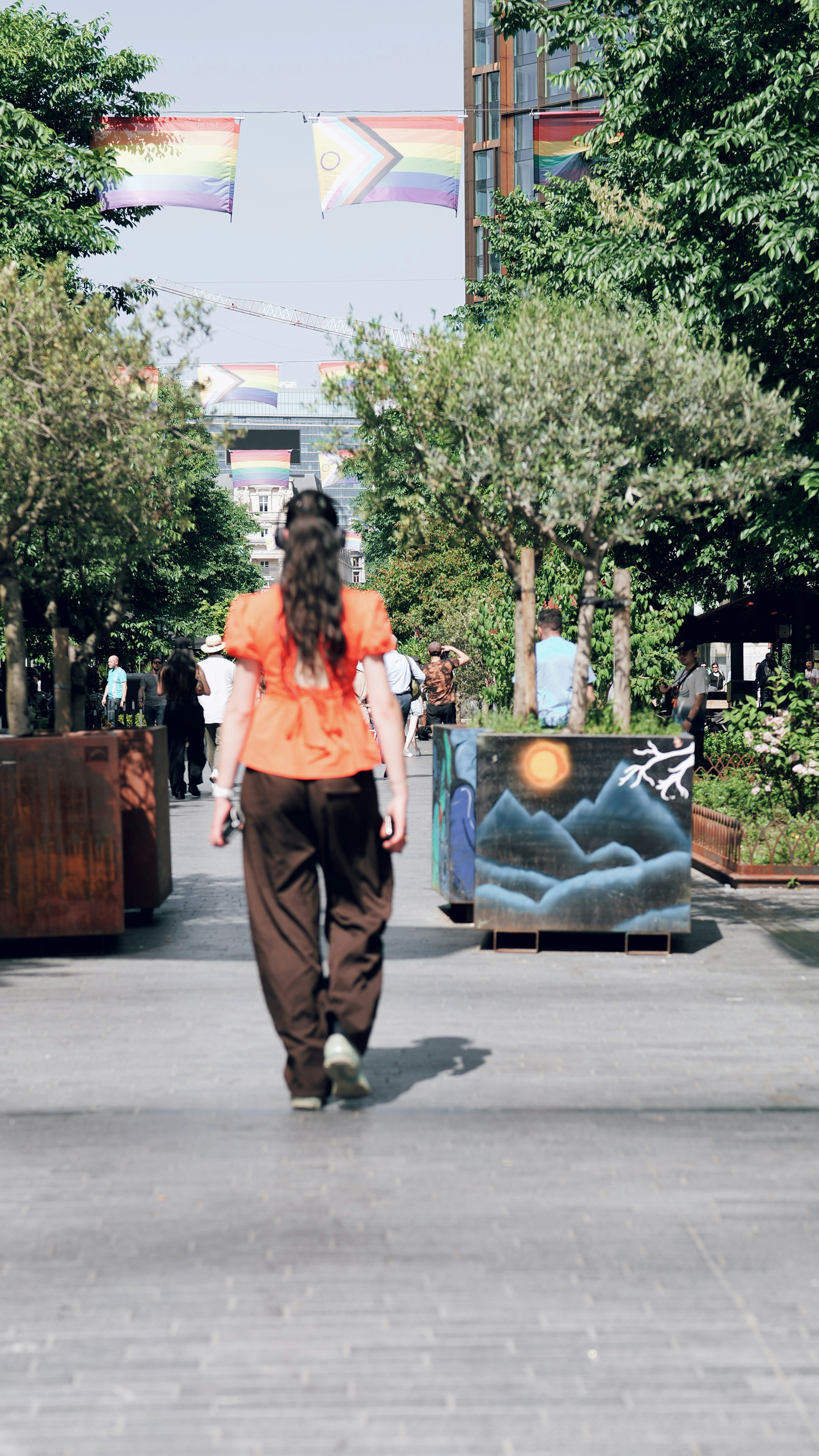 Orange-clad pedestrian strides down a sunlit urban promenade lined with rainbow banners