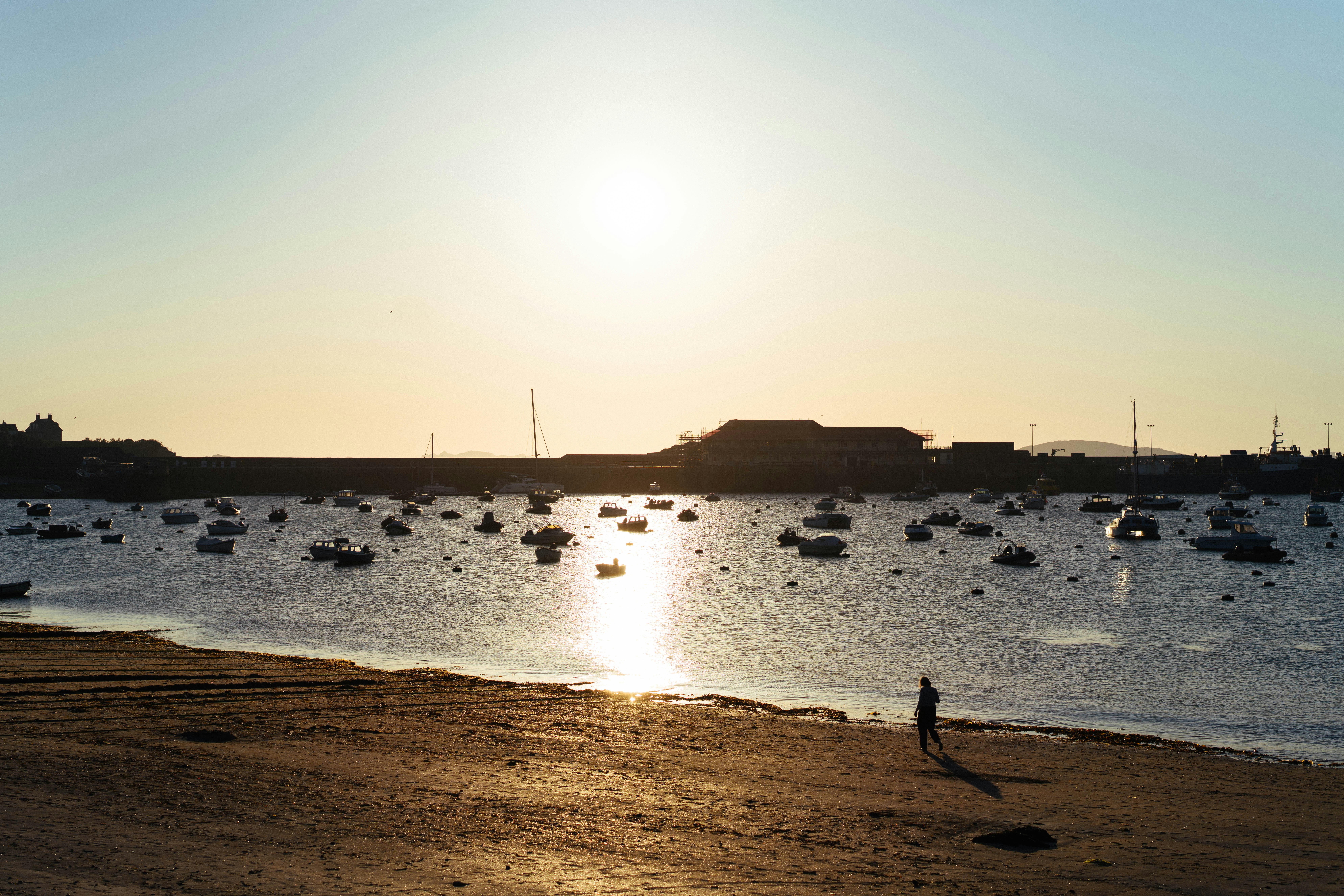 Sunlit Harbor: Boats, Water, and a lone figure on the beach at golden hour