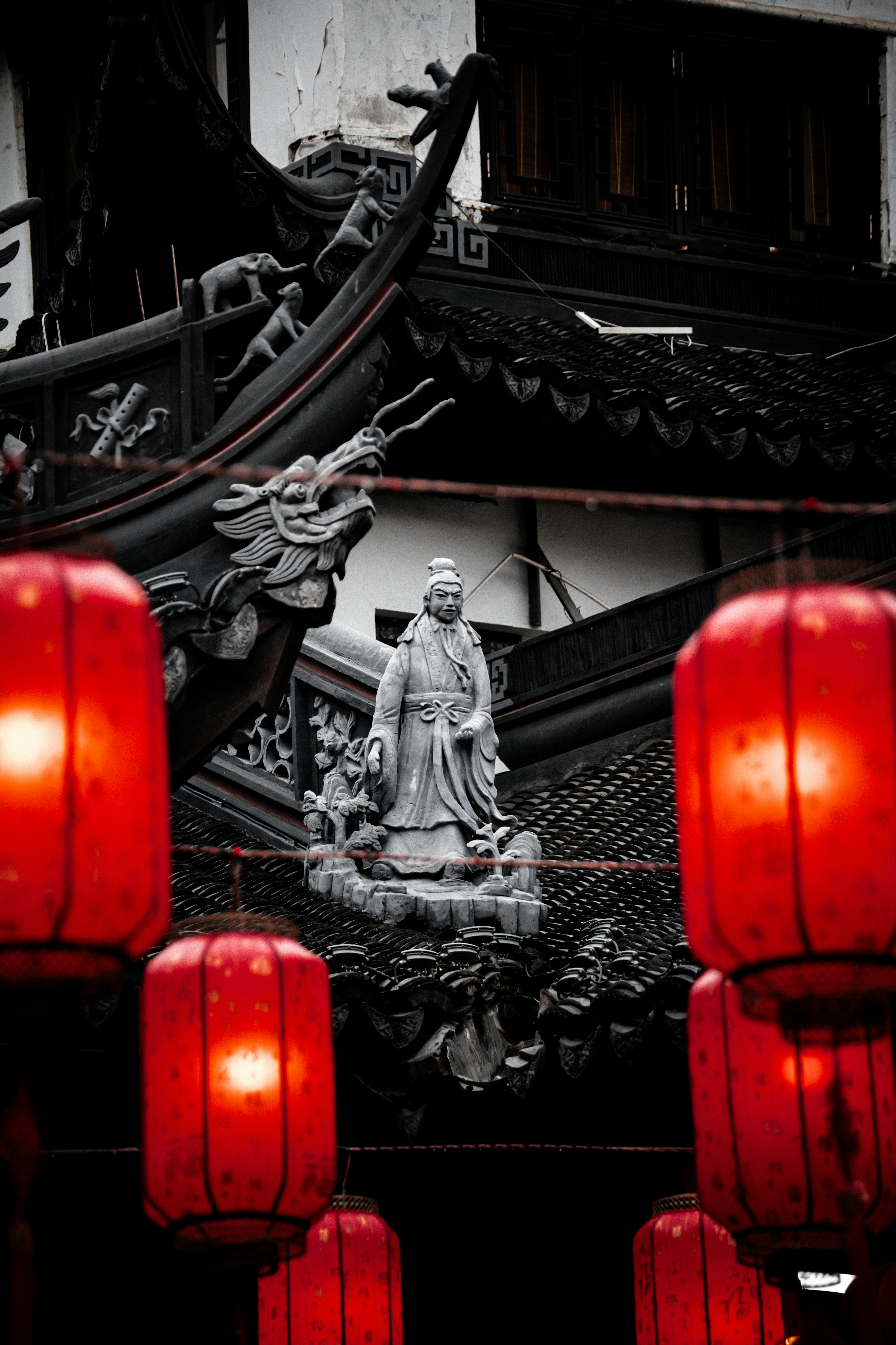 Red Lanterns framing a serene stone statue above weathered roofs