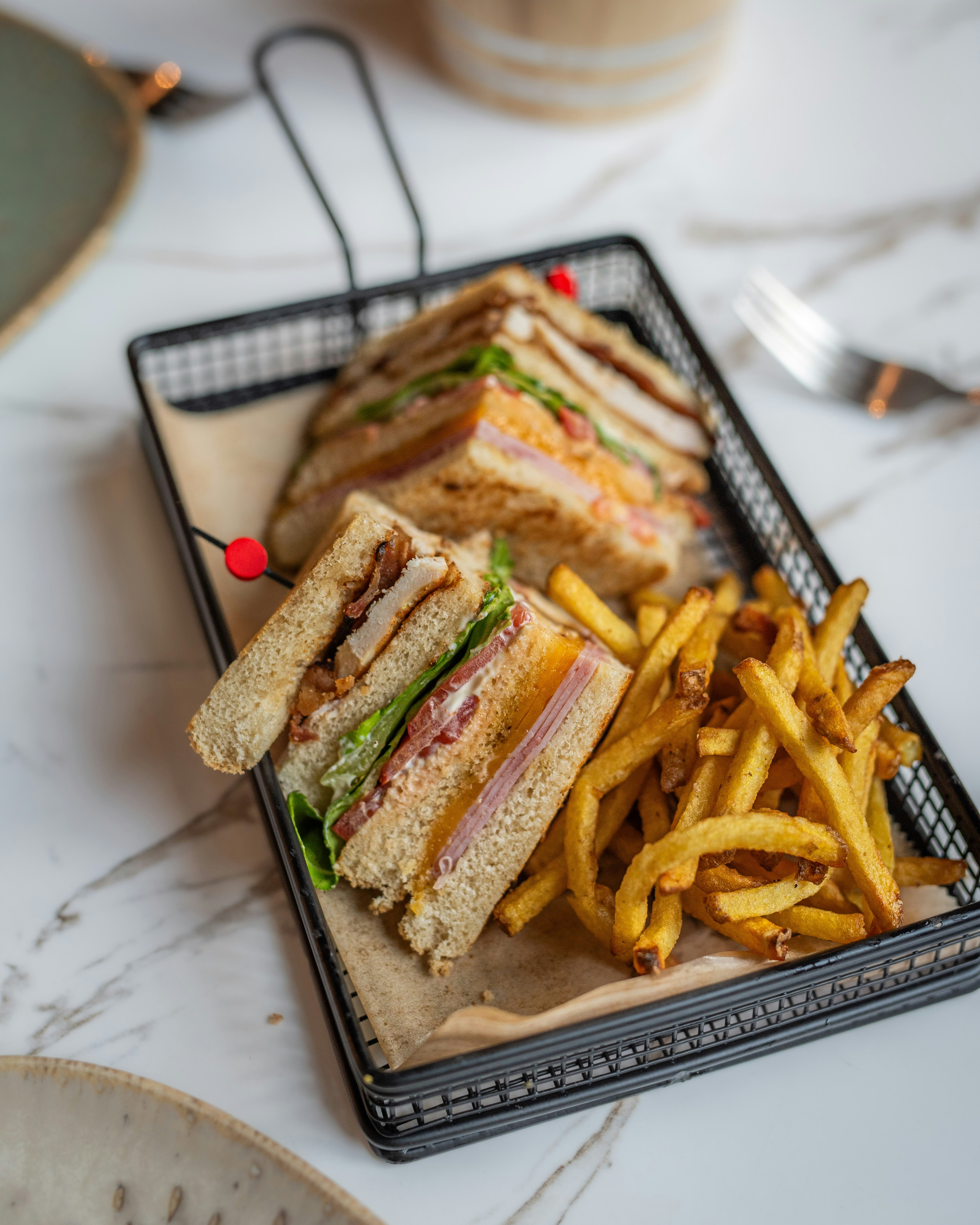 A bento-style black tray holds stacked club sandwiches with layers of turkey, bacon, lettuce, and tomato beside a pile of golden fries on a marble table.