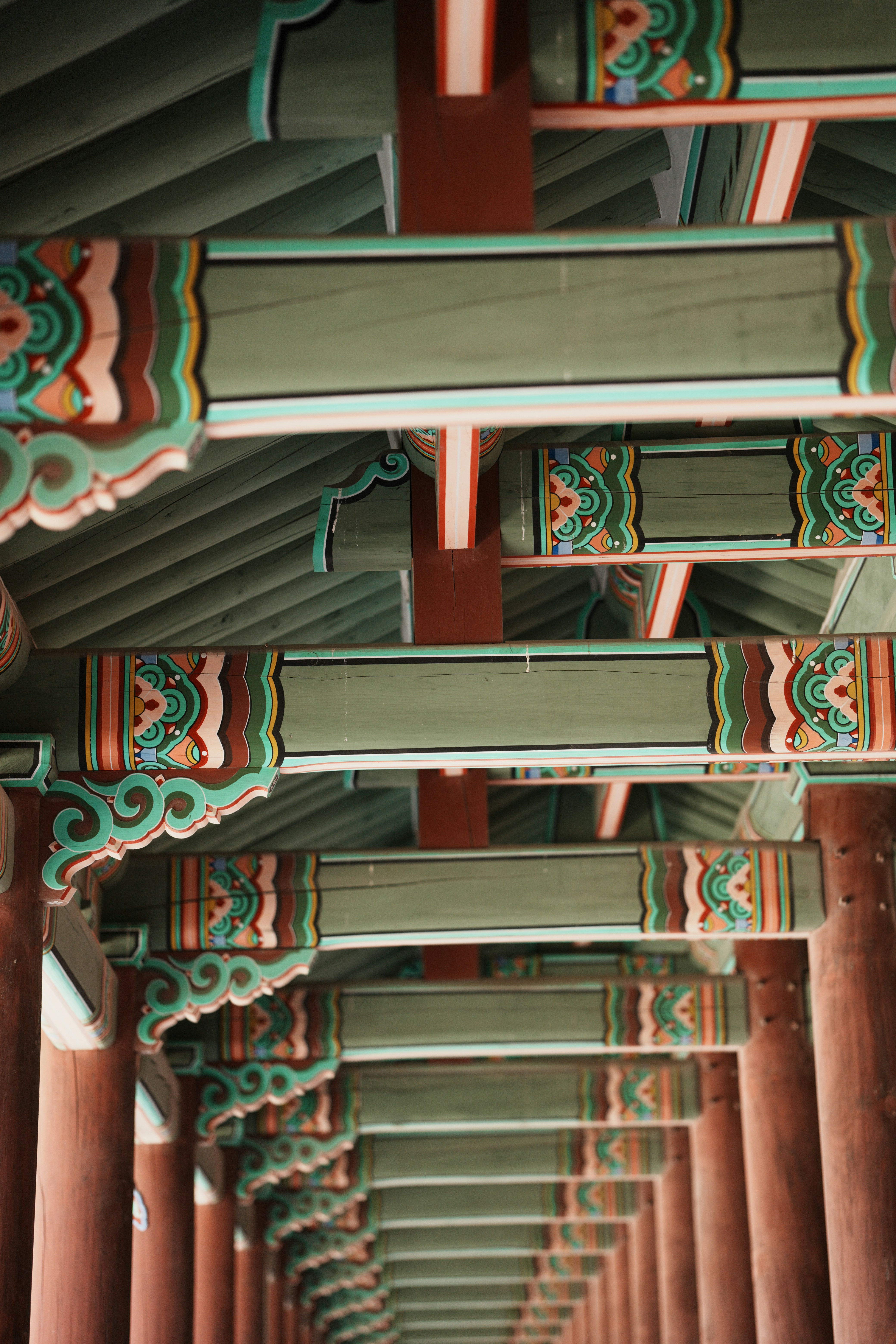 Patterned beams and columns of a traditional pavilion form a repeating architectural rhythm with vibrant green, red, and teal motifs. The image captures a symmetrical corridor of painted rafters and pillars, emphasizing depth and texture.