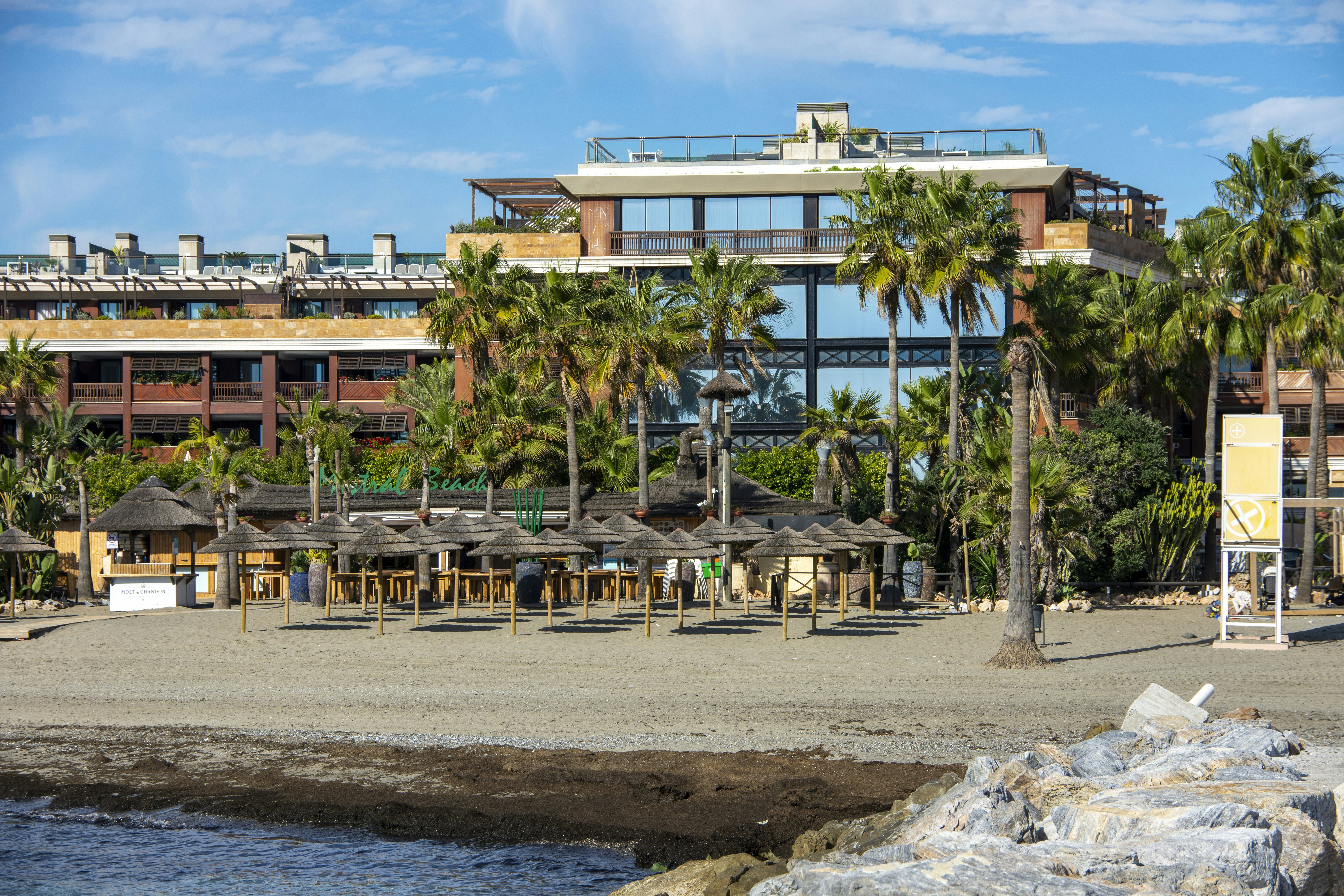 Sunlit beachfront hotel with rows of cabanas along the sandy shore, calm water lapping the beach, and a modern, glass-fronted building in the background.