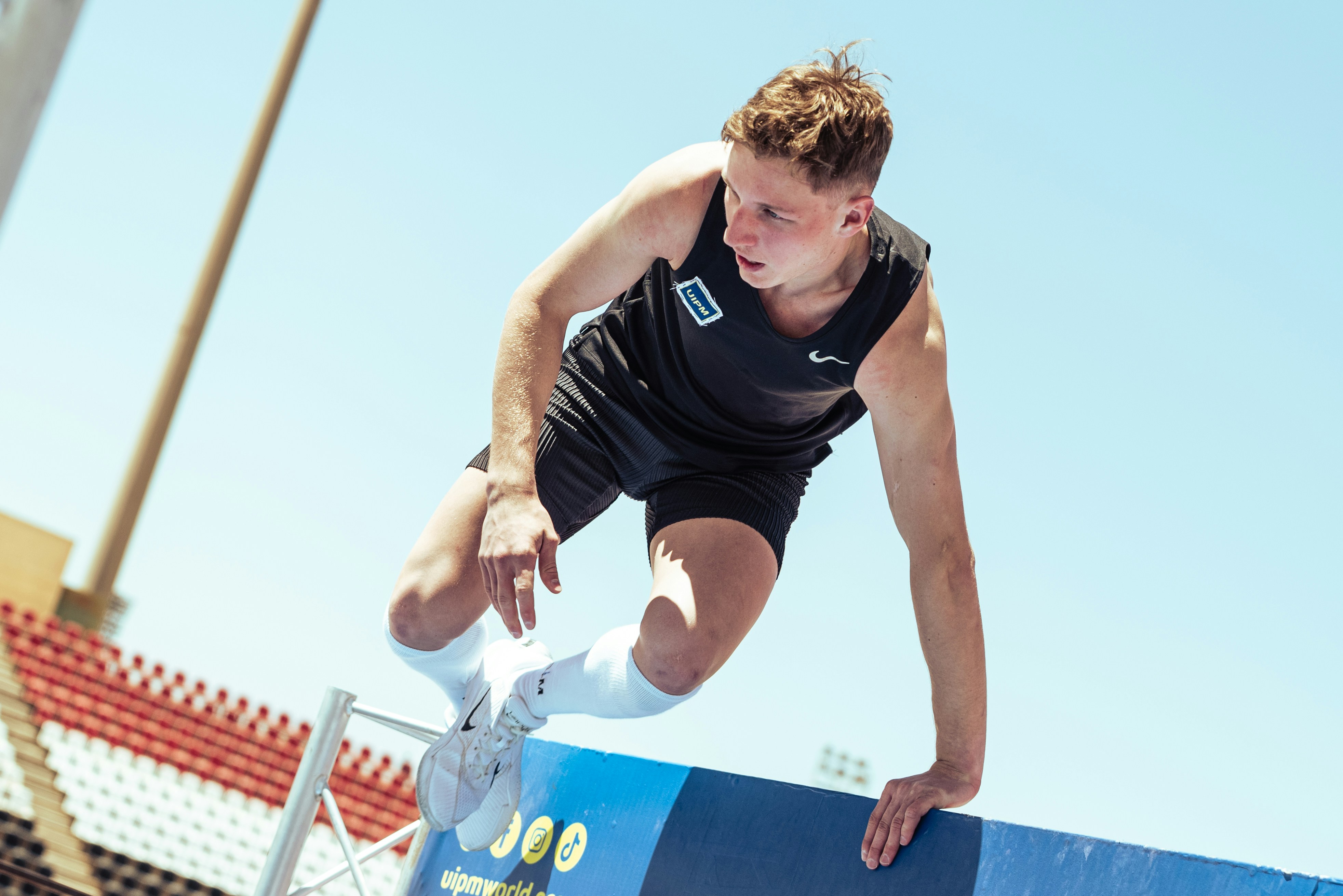 Aerial Leap: Athlete Climbing the Barrier Under a Bright Stadium Sky