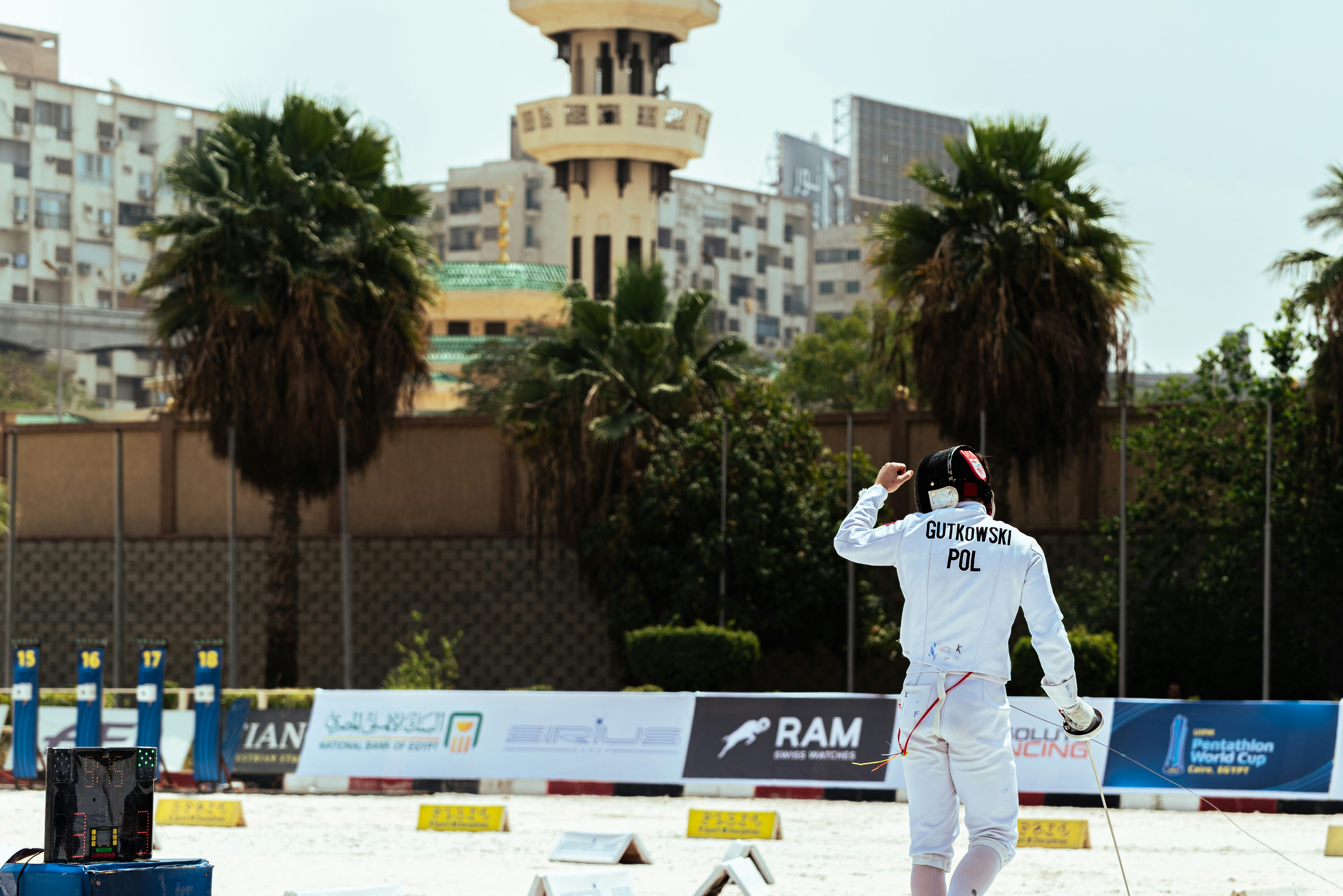 White-jersey fencer backs toward arena boundary under palm-lined backdrop