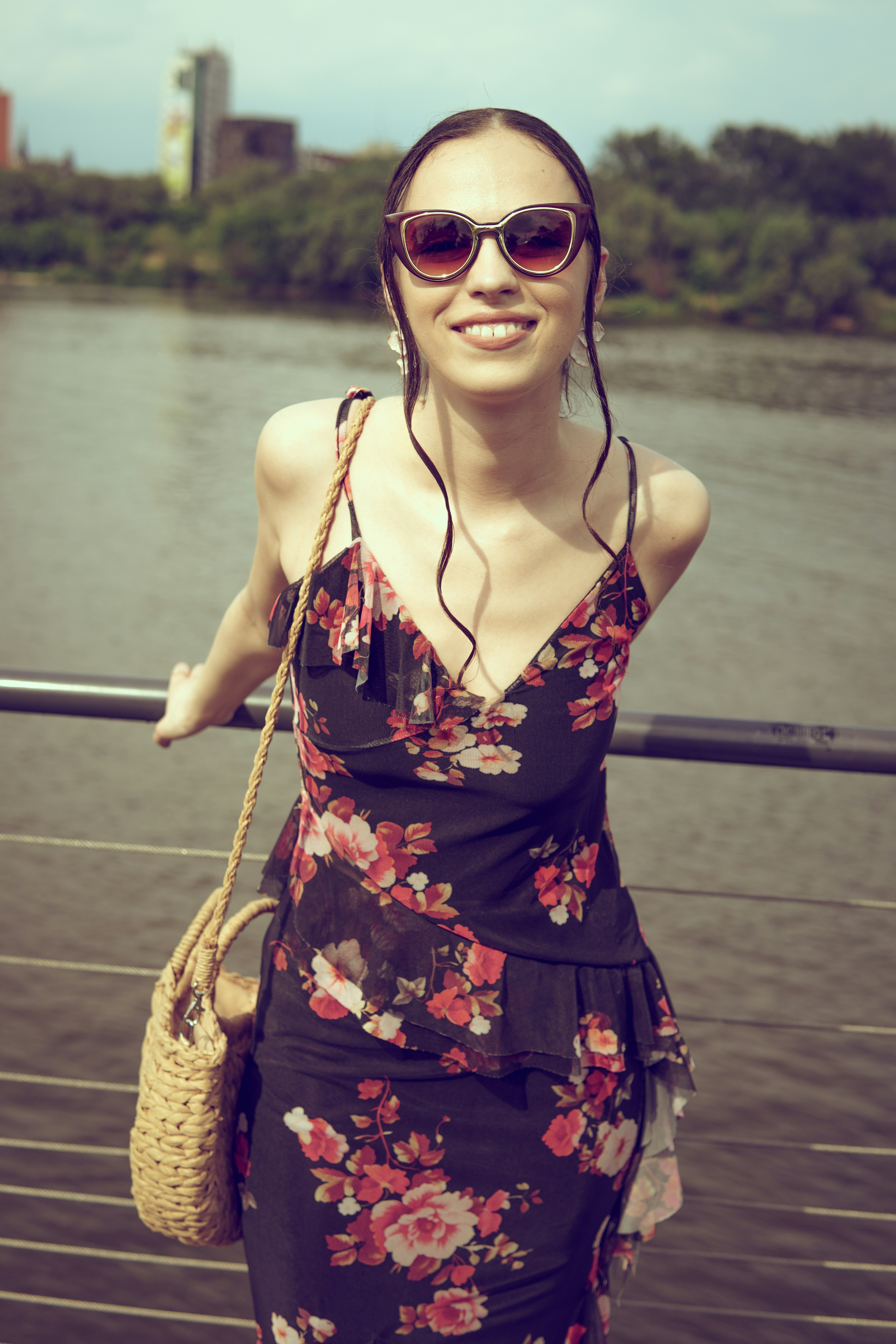 Woman in floral dress and sunglasses smiles by river.