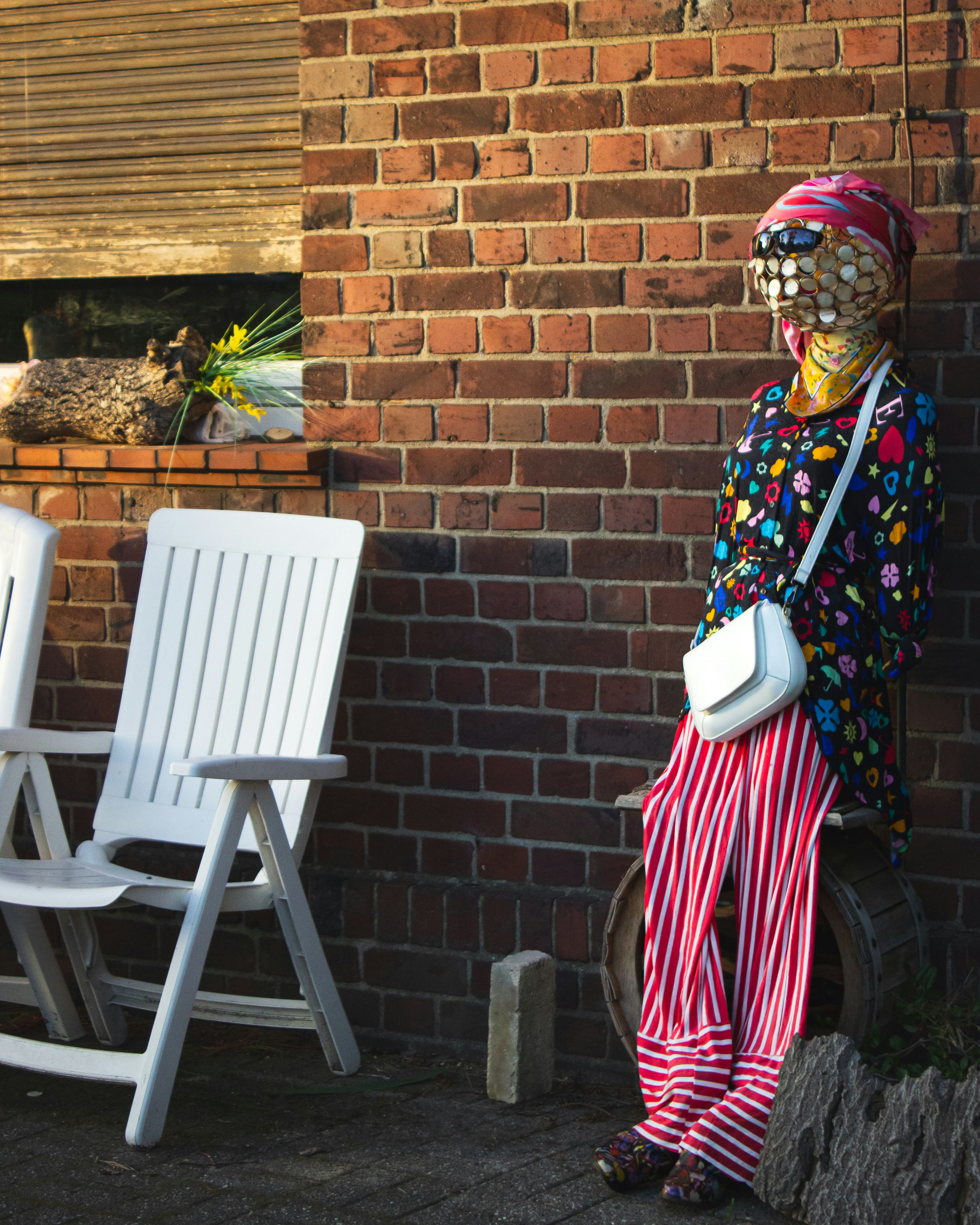 Vividly dressed mannequin-like figure leaning beside a brick wall; white chairs sit nearby, sunlight highlighting textures and colors.