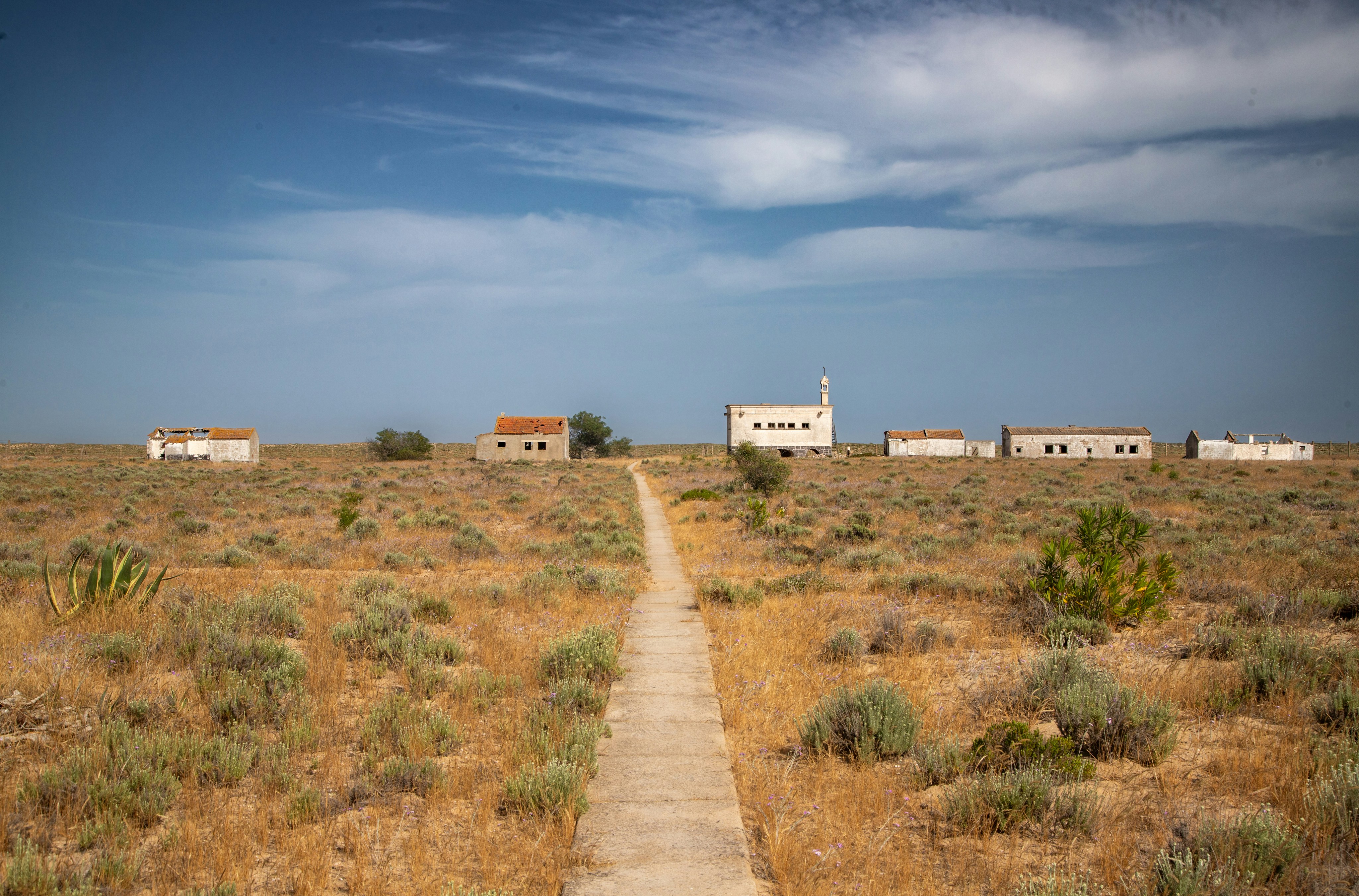Dusty corridor leads to a row of weathered buildings under a wide blue sky in an arid landscape.