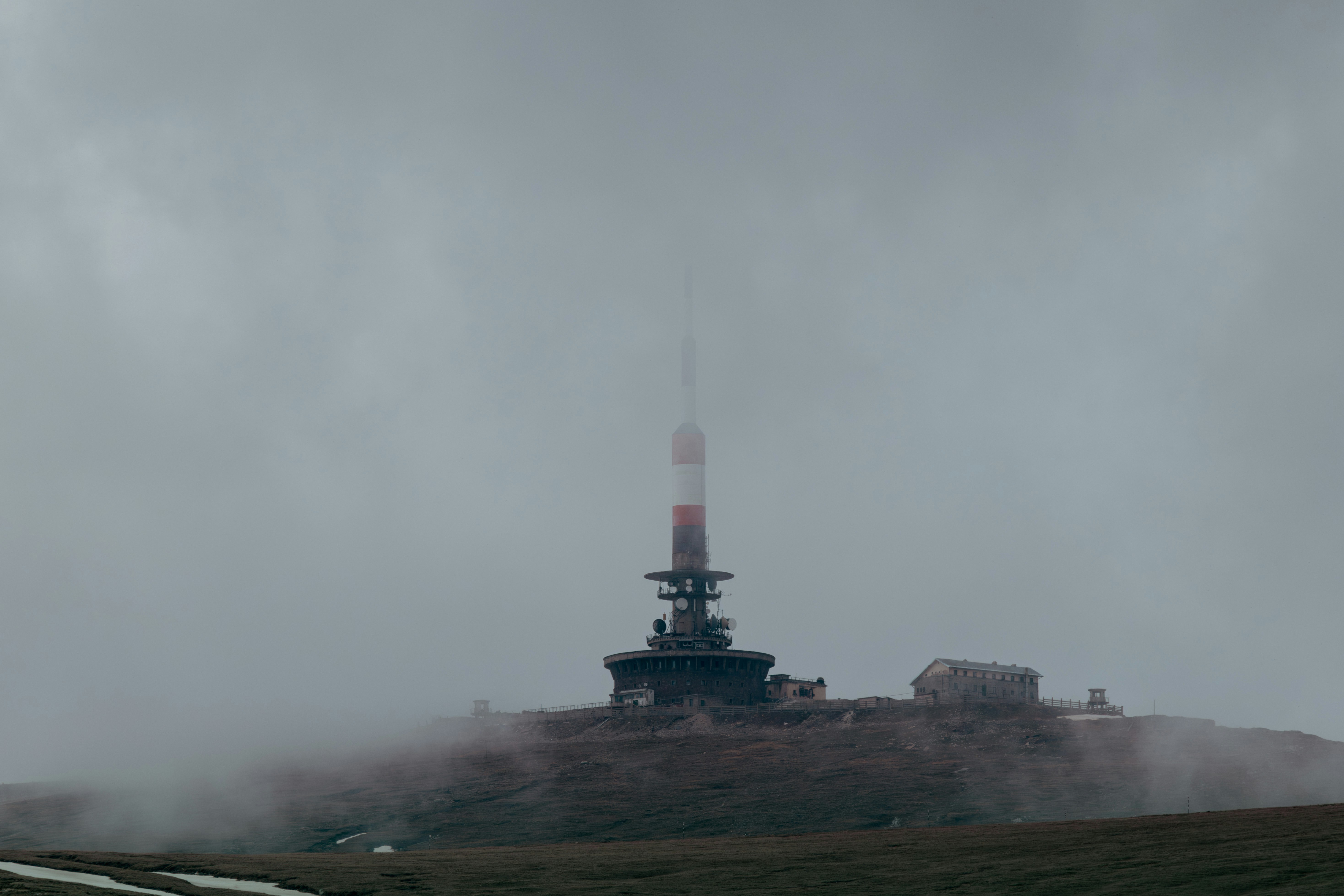 Fog wraps a barren hill where a solitary transmission tower rises, with a weathered building nearby; the scene emphasizes isolation and moody atmosphere. This is a photographic capture, not a painting or illustration.