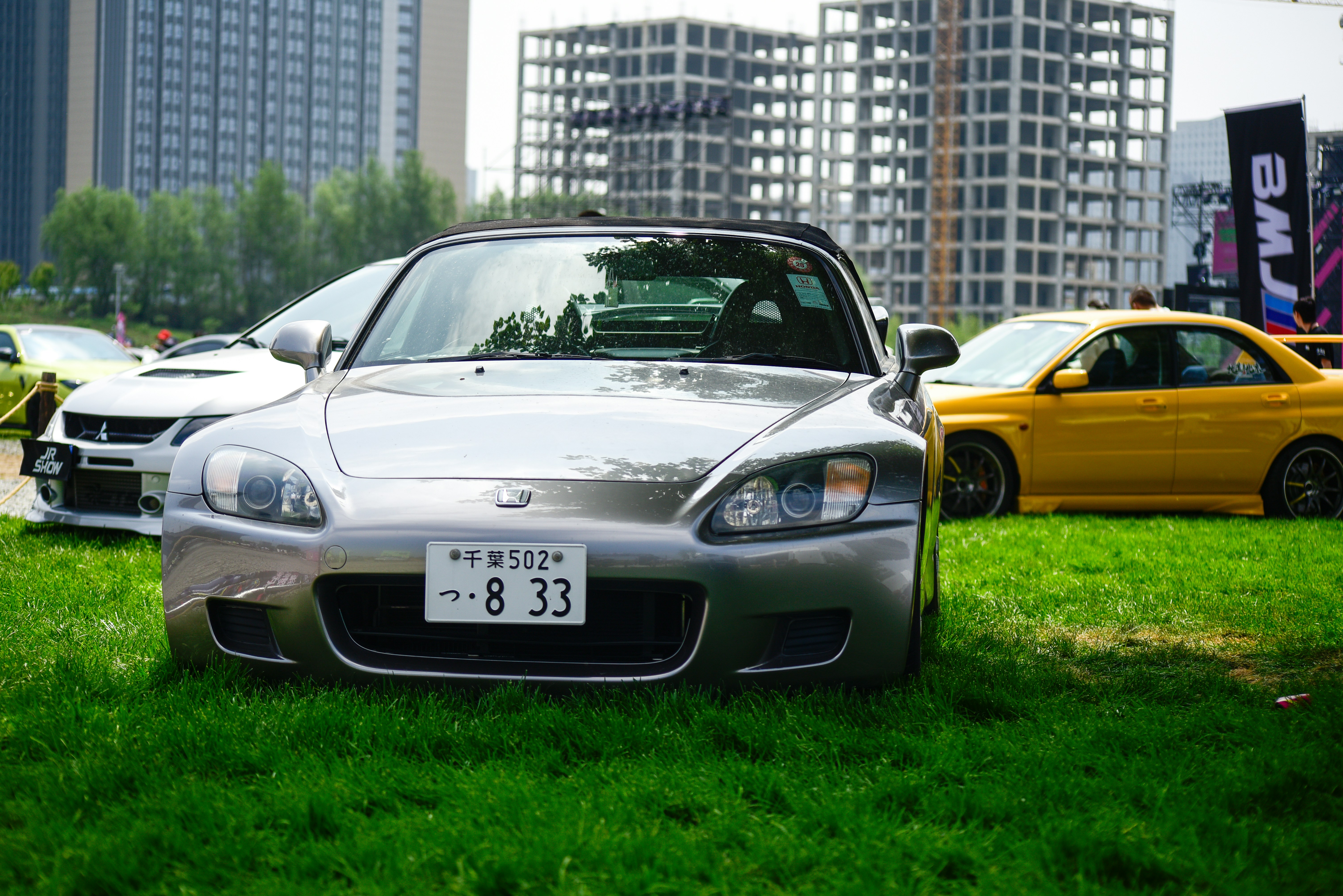 Silver MX-5 at a grassy overlook under urban vigilante towers