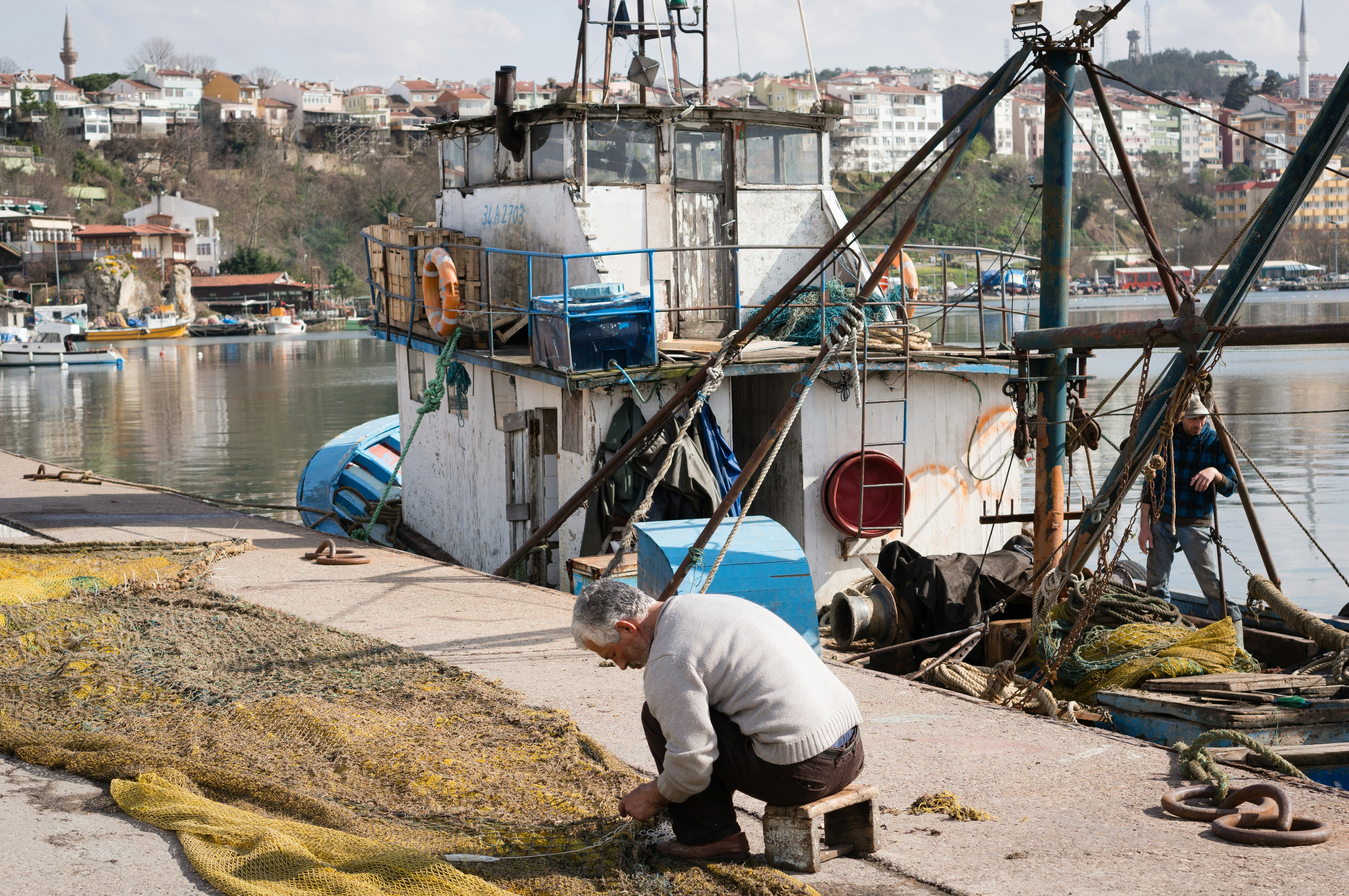 Weathered fisher at the quay: nets, boat, and distant hillside town
