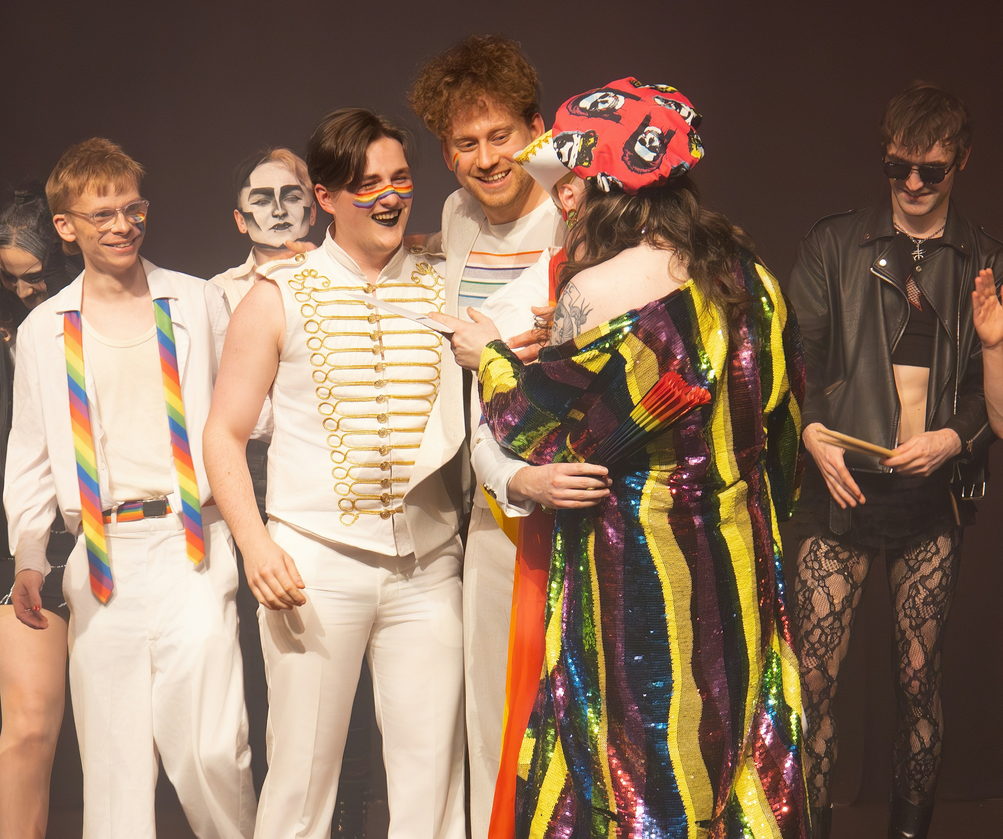 Diverse group of performers in vibrant costumes embracing on stage.