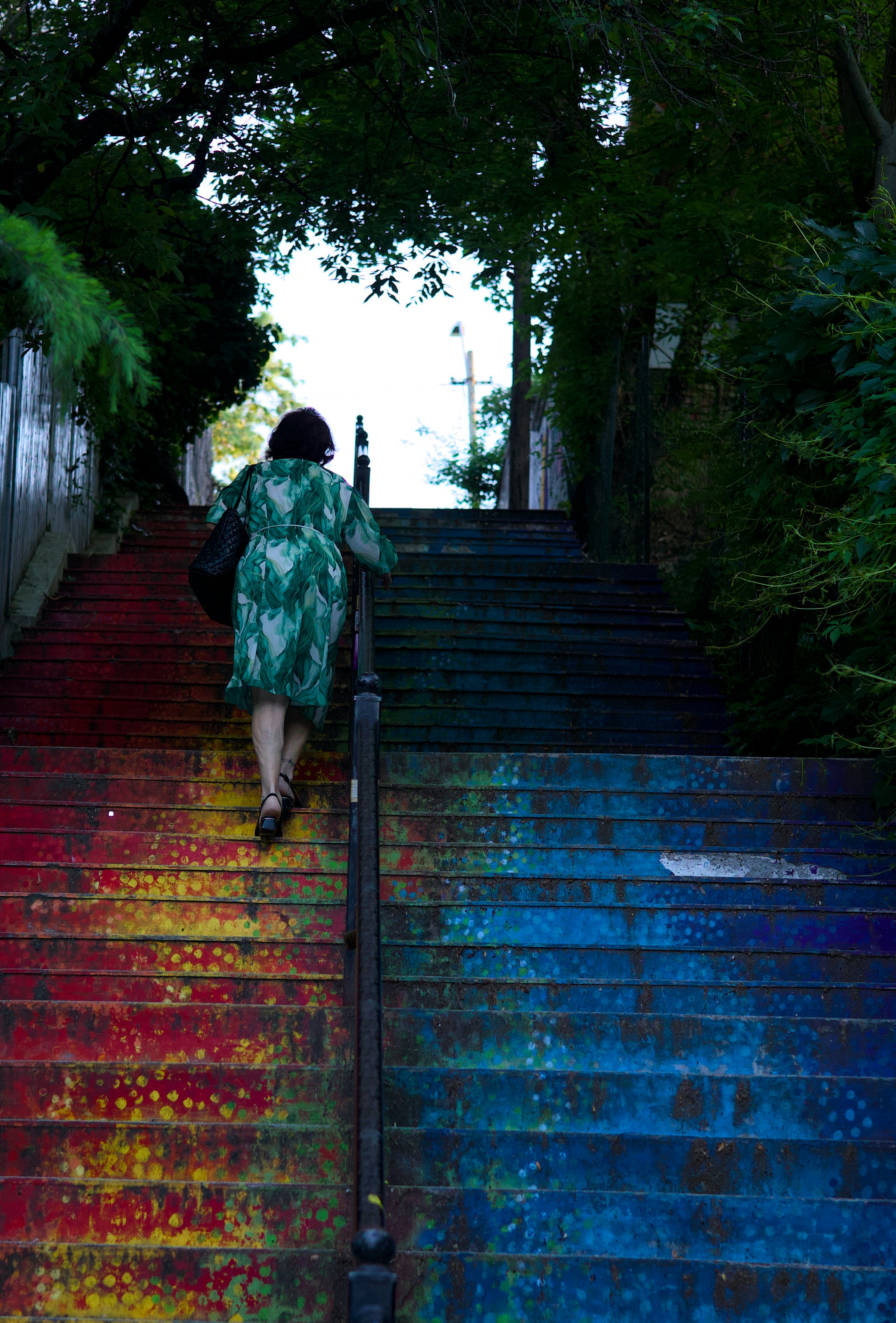 Colorful ascent: a solitary traveller climbs rainbow-painted steps under a leafy arch