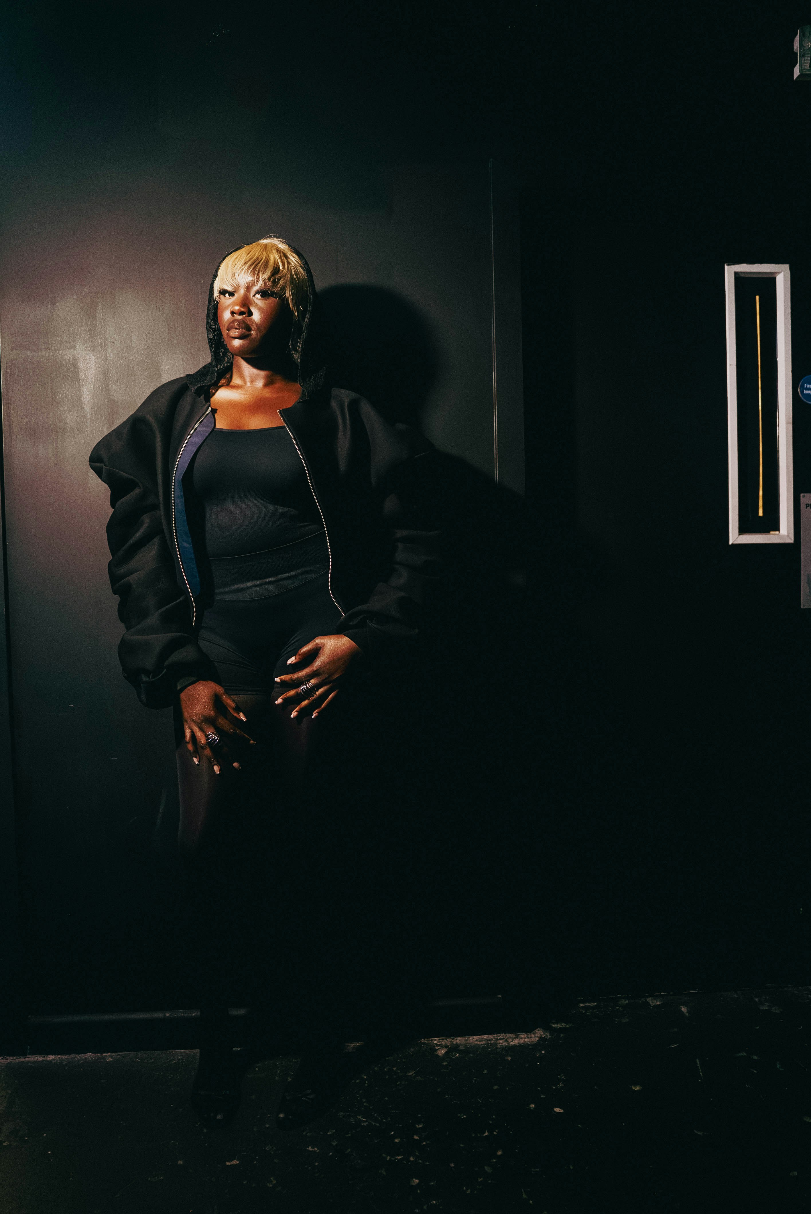 Woman with blonde hair in black jacket against dark wall.