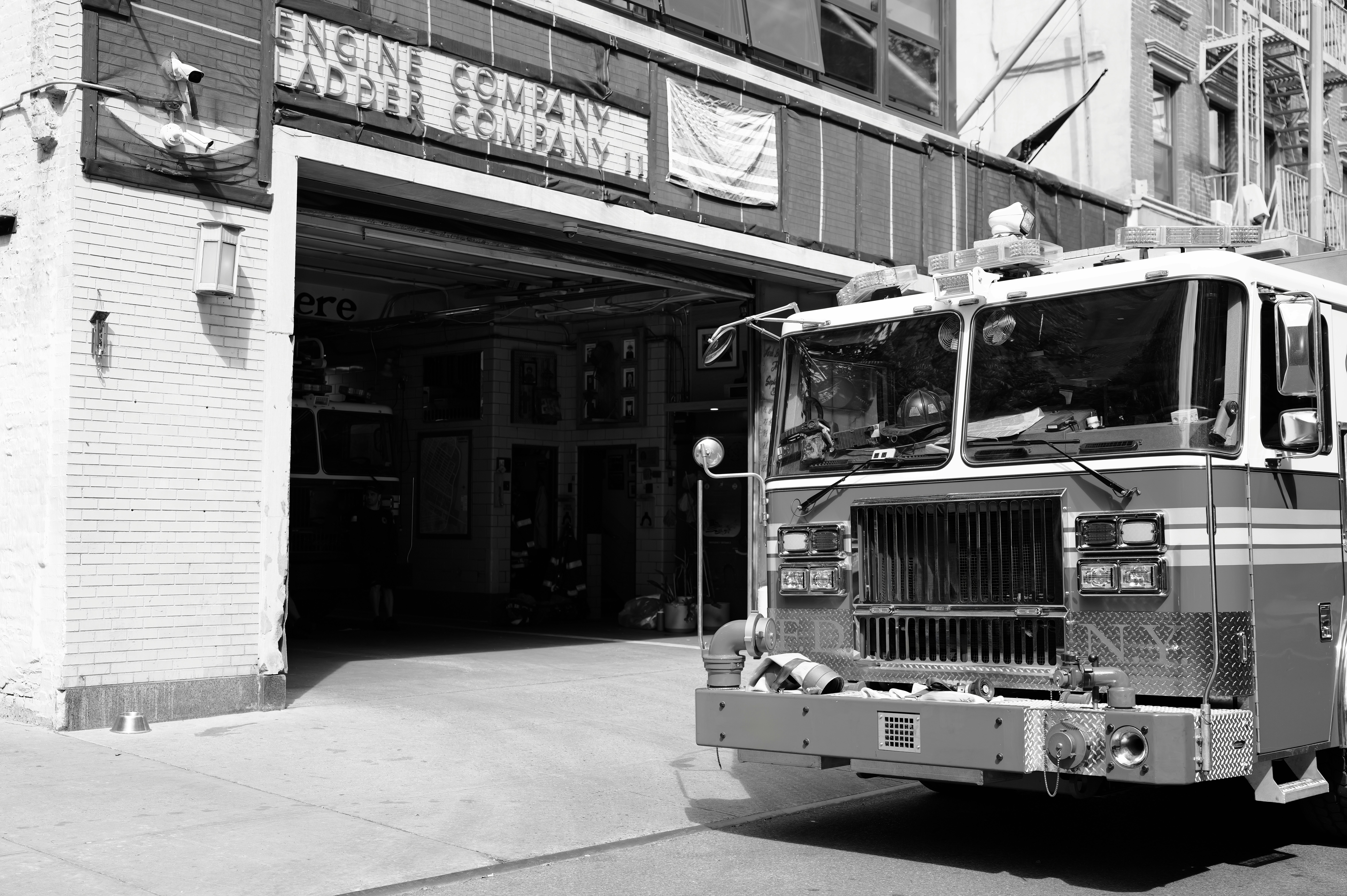 Black-and-white scene of a fire engine parked at a station entrance with an open bay, highlighting the vehicle and urban firehouse context.