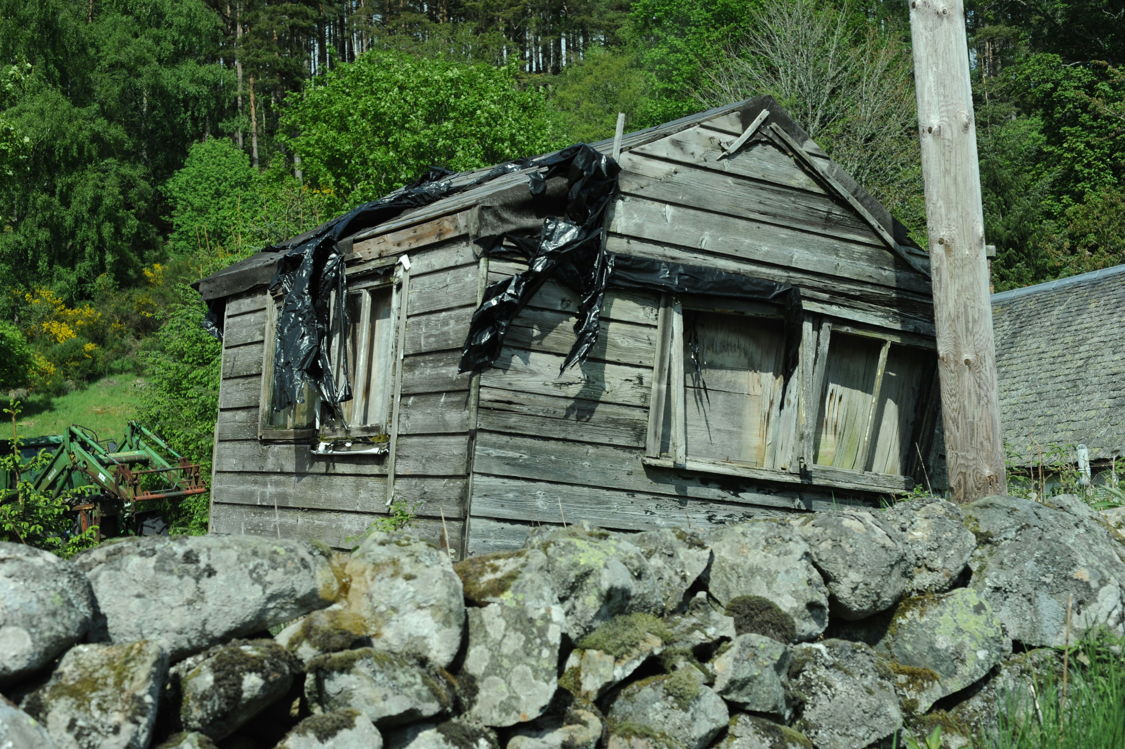 Rundown wooden shack leaning on a stone wall amid greenery, with a damaged roof and weathered planks. A rustic, weather-beaten structure emphasizing decay.