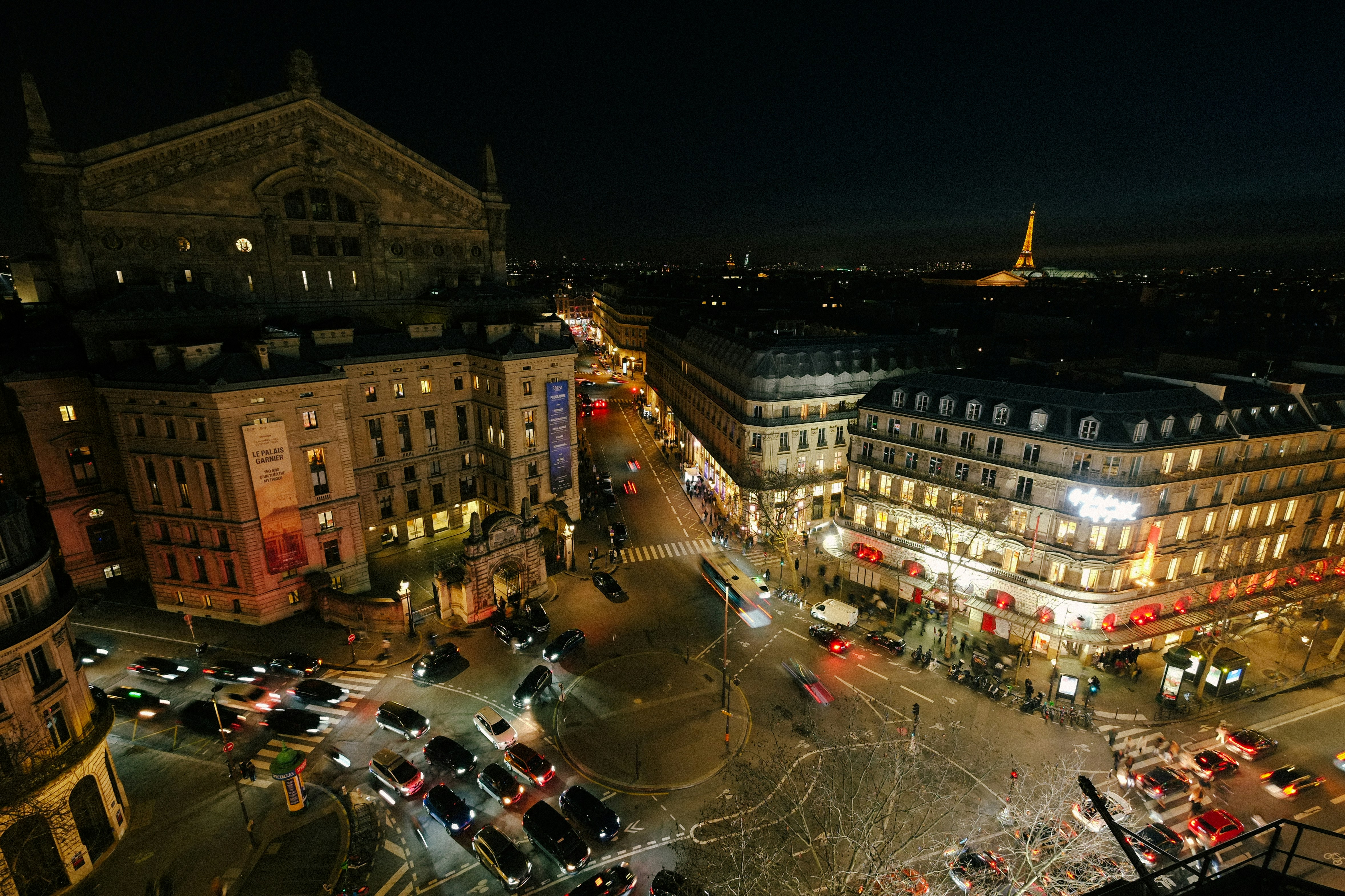 Nighttime cityscape of a bustling European square with illuminated buildings and a distant illuminated tower, captured from a high vantage point. This is a wide urban scene showing motion and light trails.