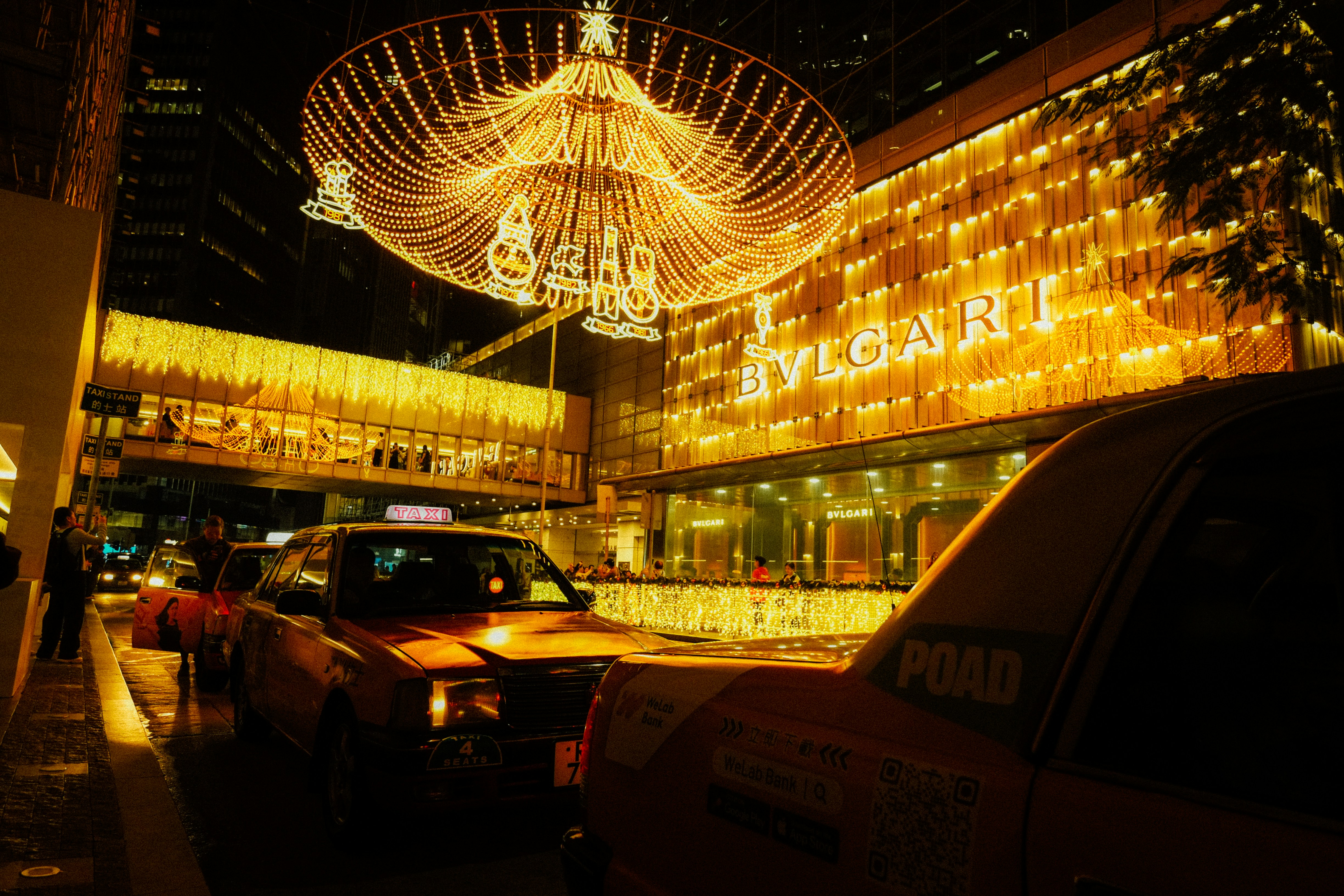 Golden-lit storefronts blaze with luxury signage and a chandelier-like overhead illumination, cars line the street, capturing a busy urban nightlife scene.