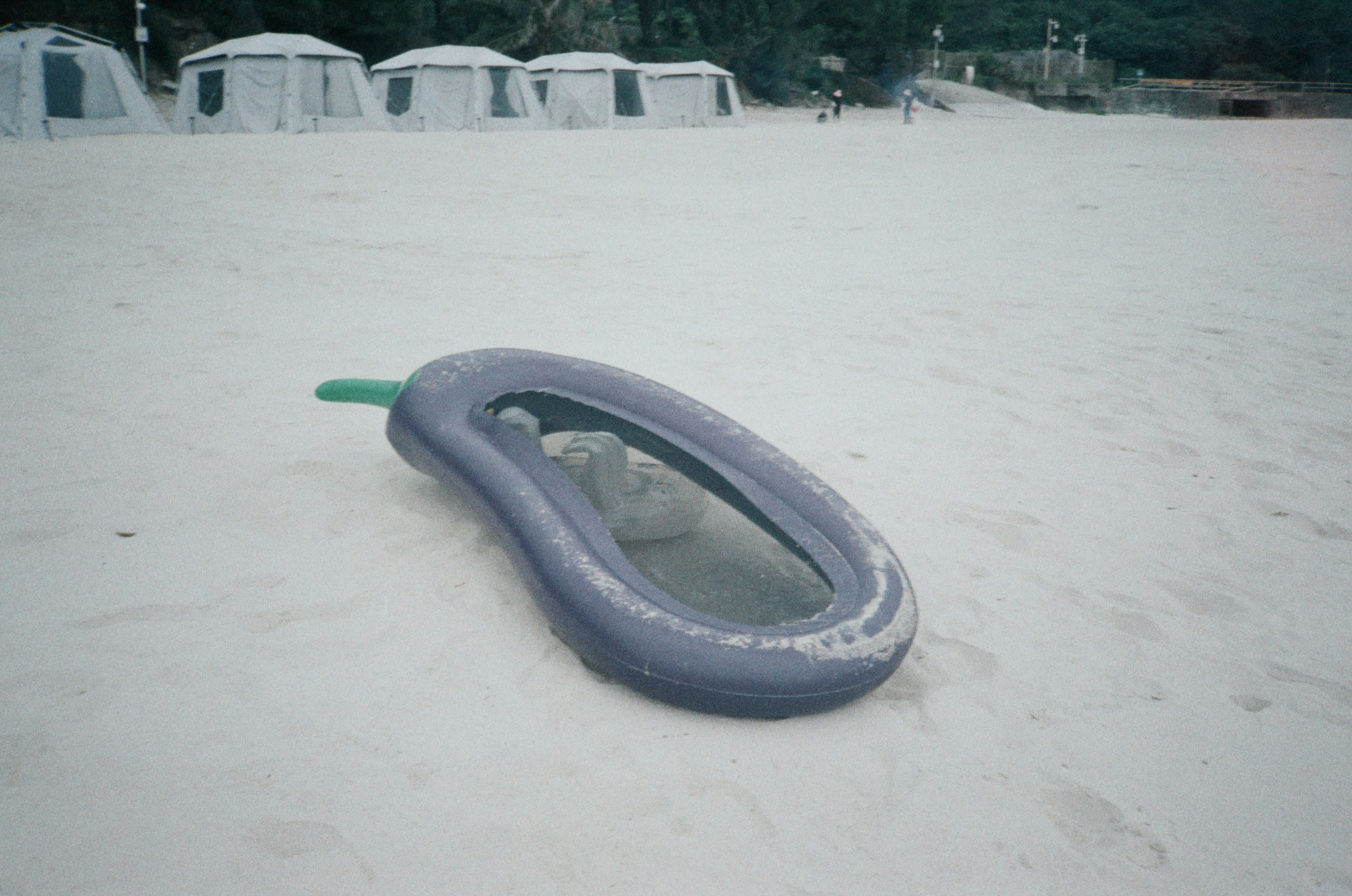 Weathered purple flotation ring rests on sunlit beach with distant tents