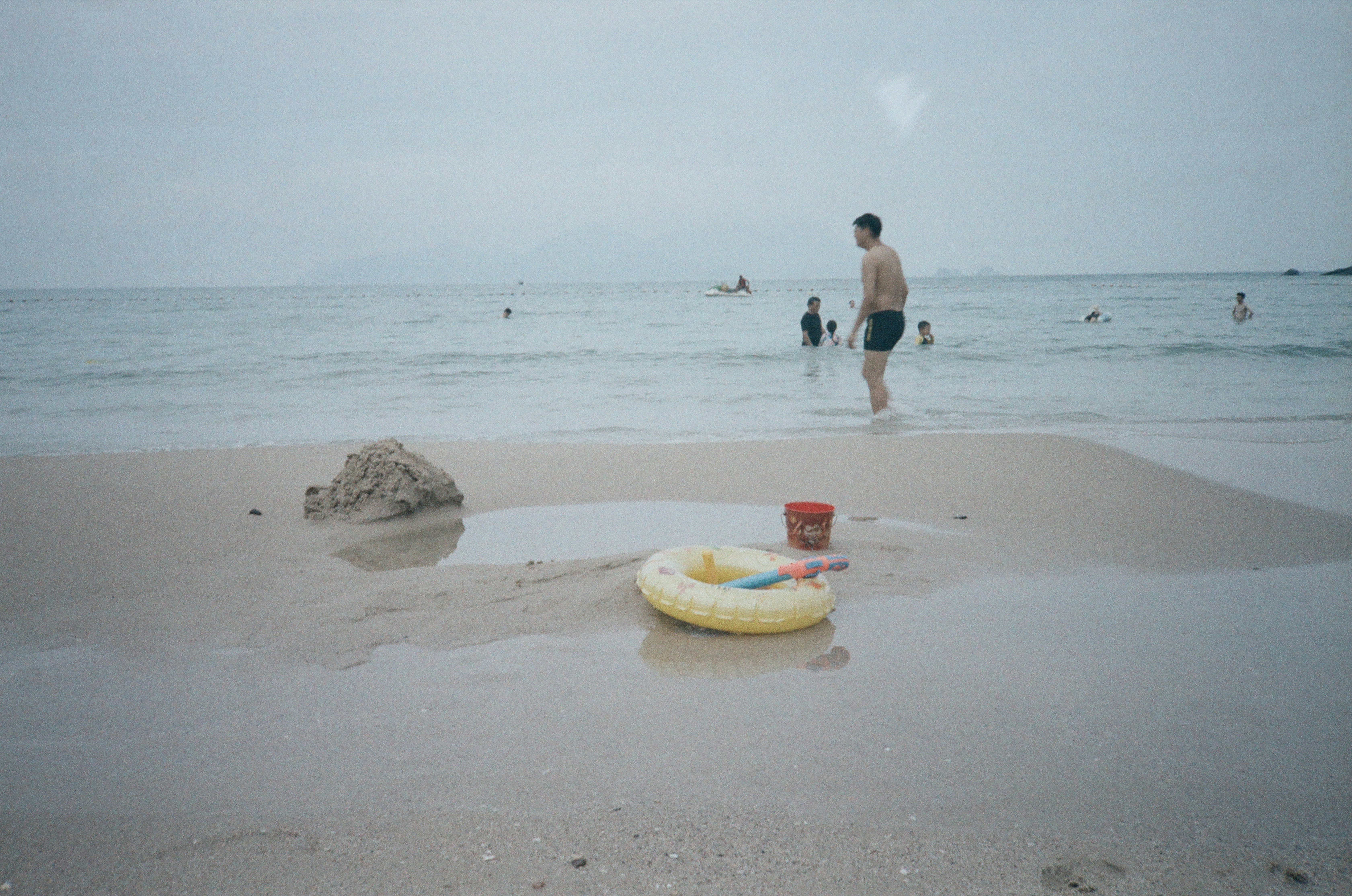 Quiet beach with a small inflatable ring and sand toys in the foreground; people wading and swimming near the calm sea, under a pale sky.
