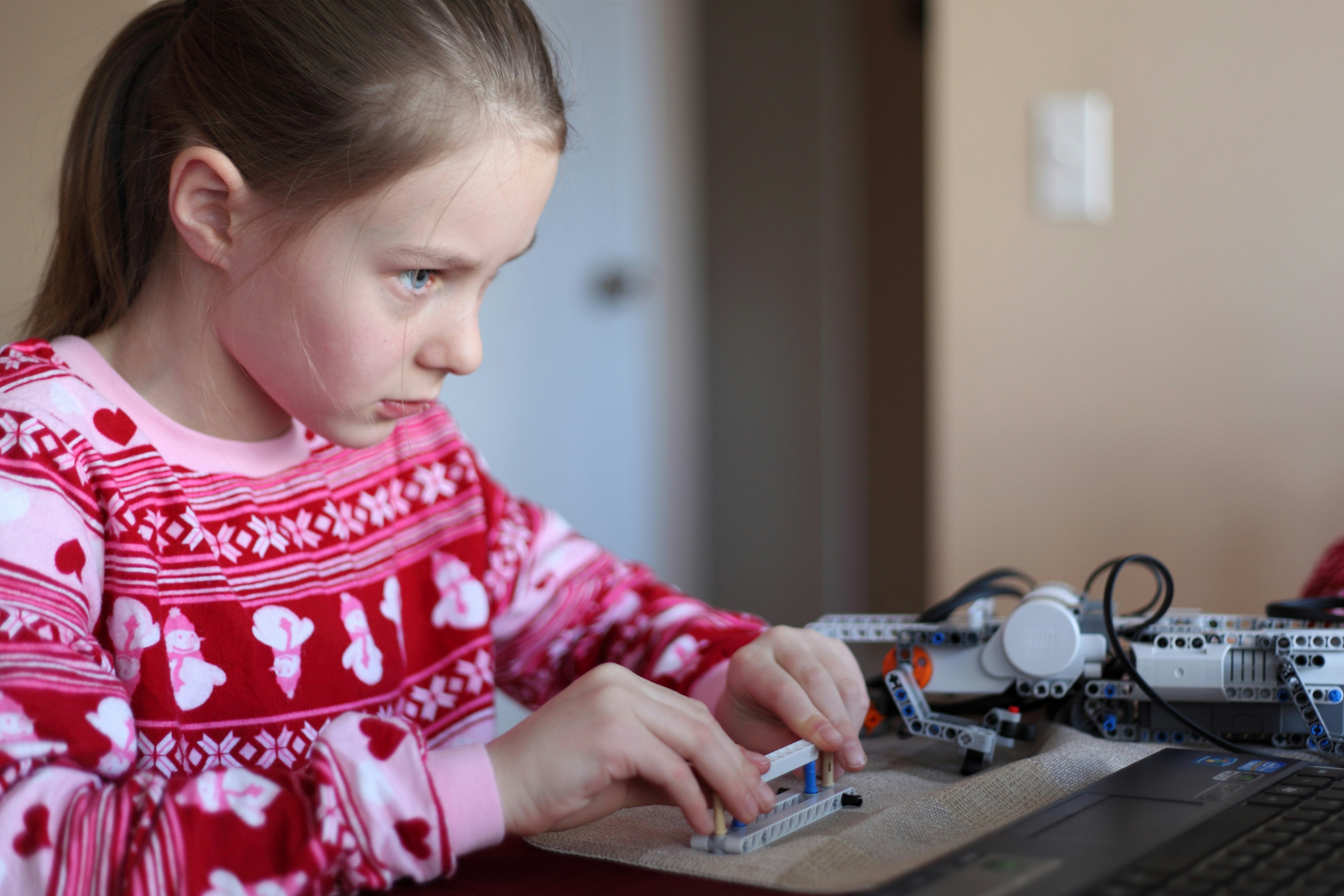 A young girl in a pink patterned sweater focuses intently on assembling a mechanical project at a desk, with a robotics kit and keyboard visible in the frame.