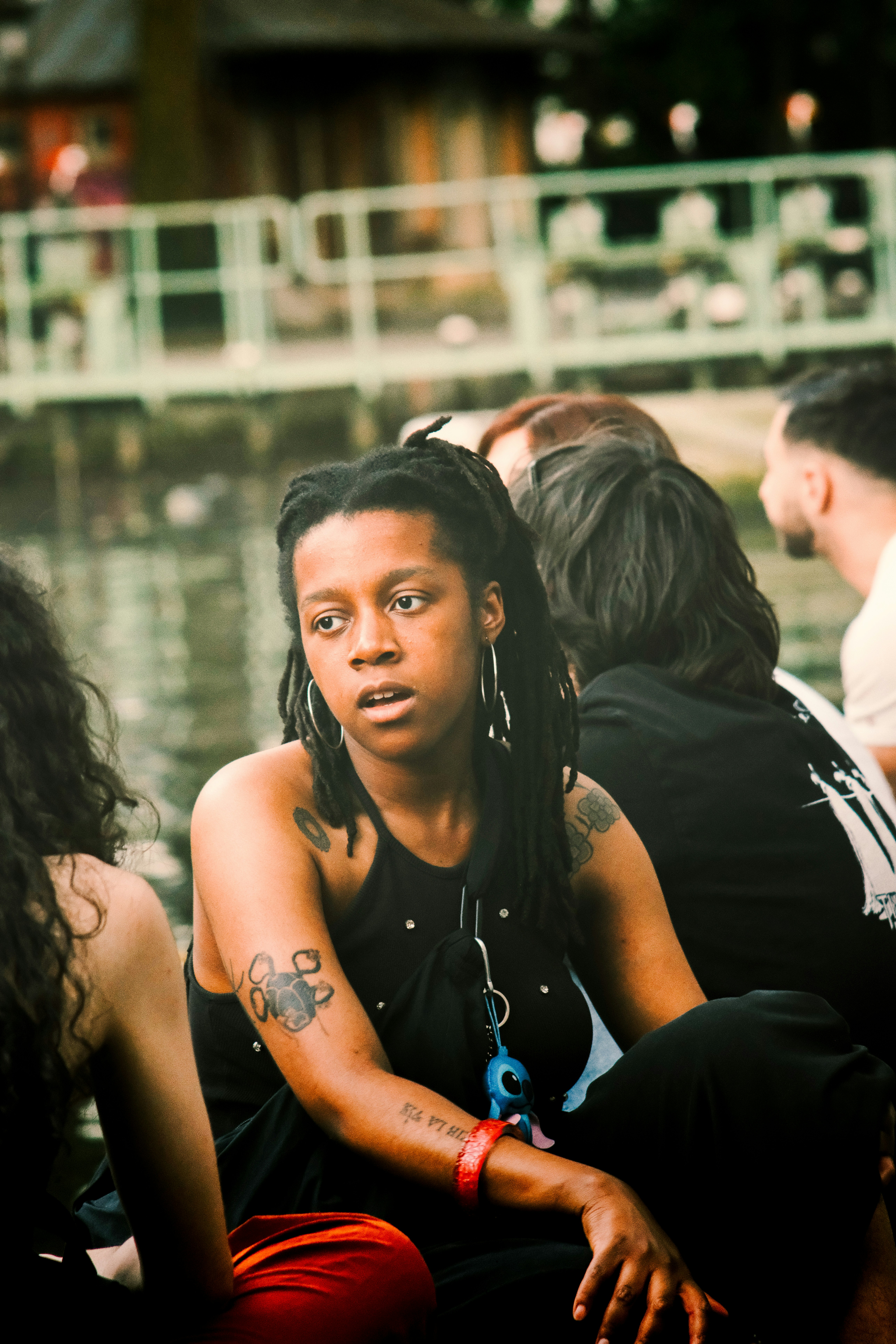A woman with dreadlocks and tattoos sitting outdoors near water.