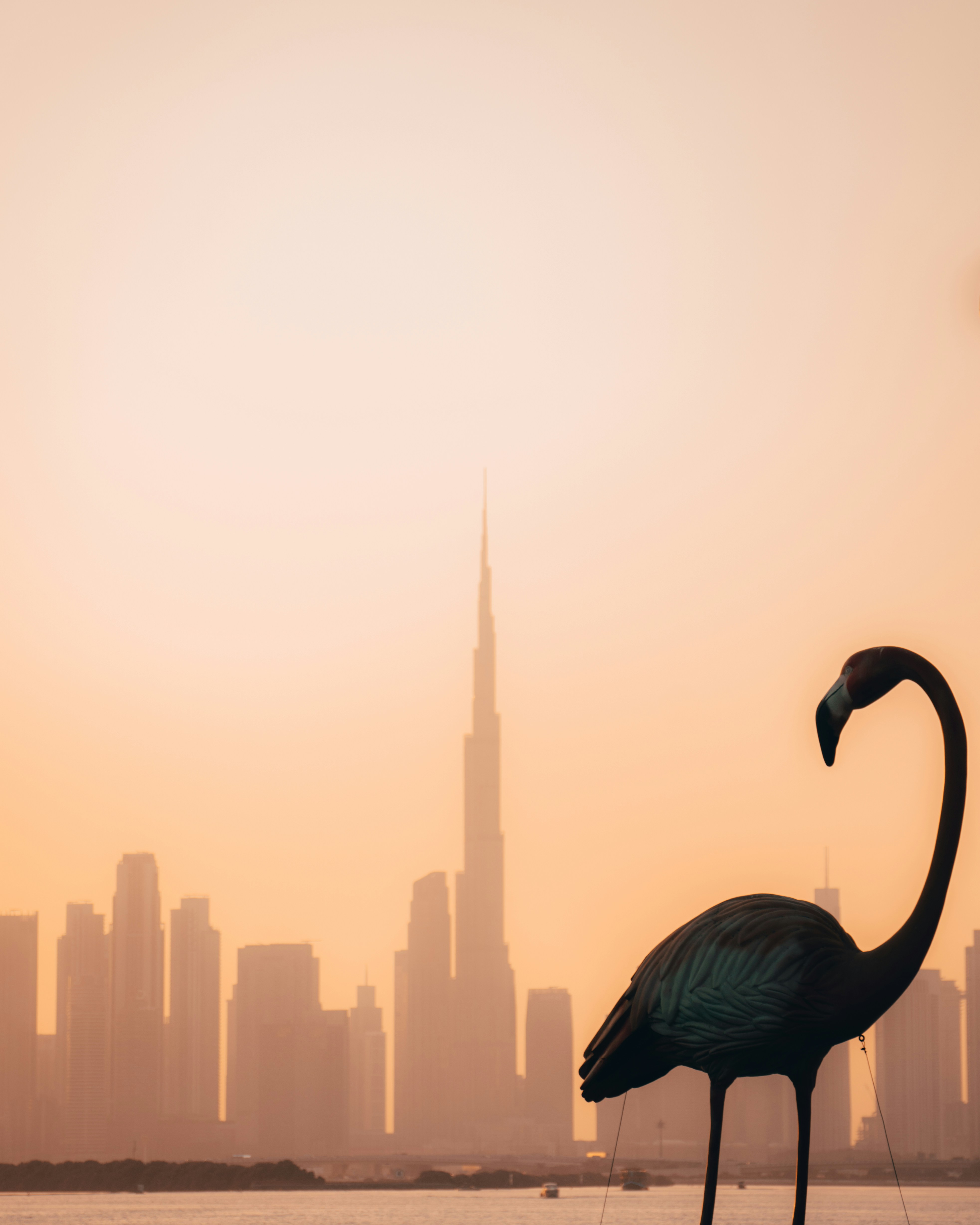 A cinematic sunset view of Dubai featuring the iconic Burj Khalifa rising through the warm haze, while a flamingo sculpture stands in the foreground overlooking the calm waters. The soft golden tones and minimalist composition create a peaceful, dreamy atmosphere that captures the beauty and elegance of the city at dusk.