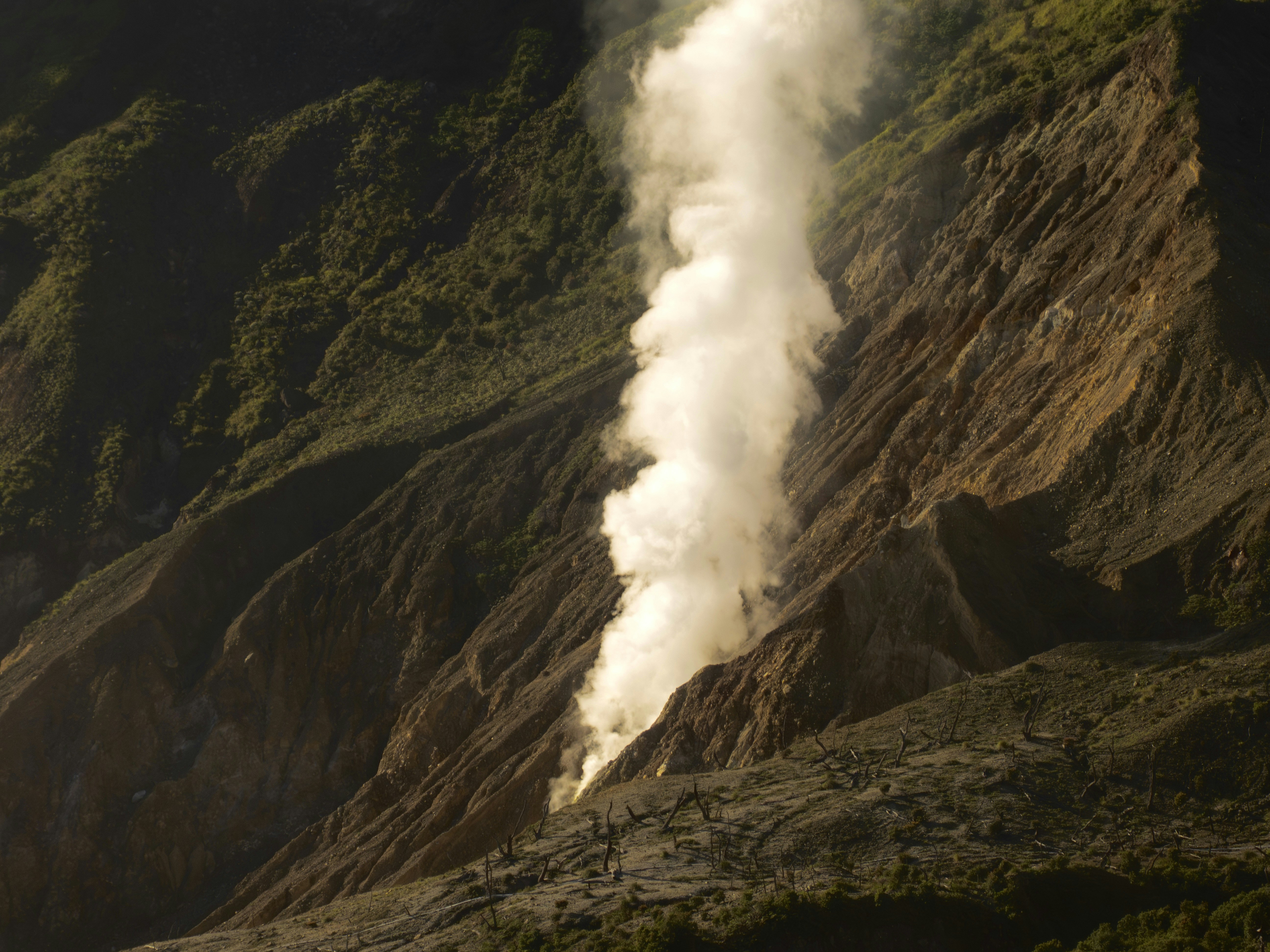 Steam plume rises from a rugged volcano hillside, captured with a landscape orientation to emphasize the jagged terrain and natural steam. Live texture and contrast highlight the geologic details.