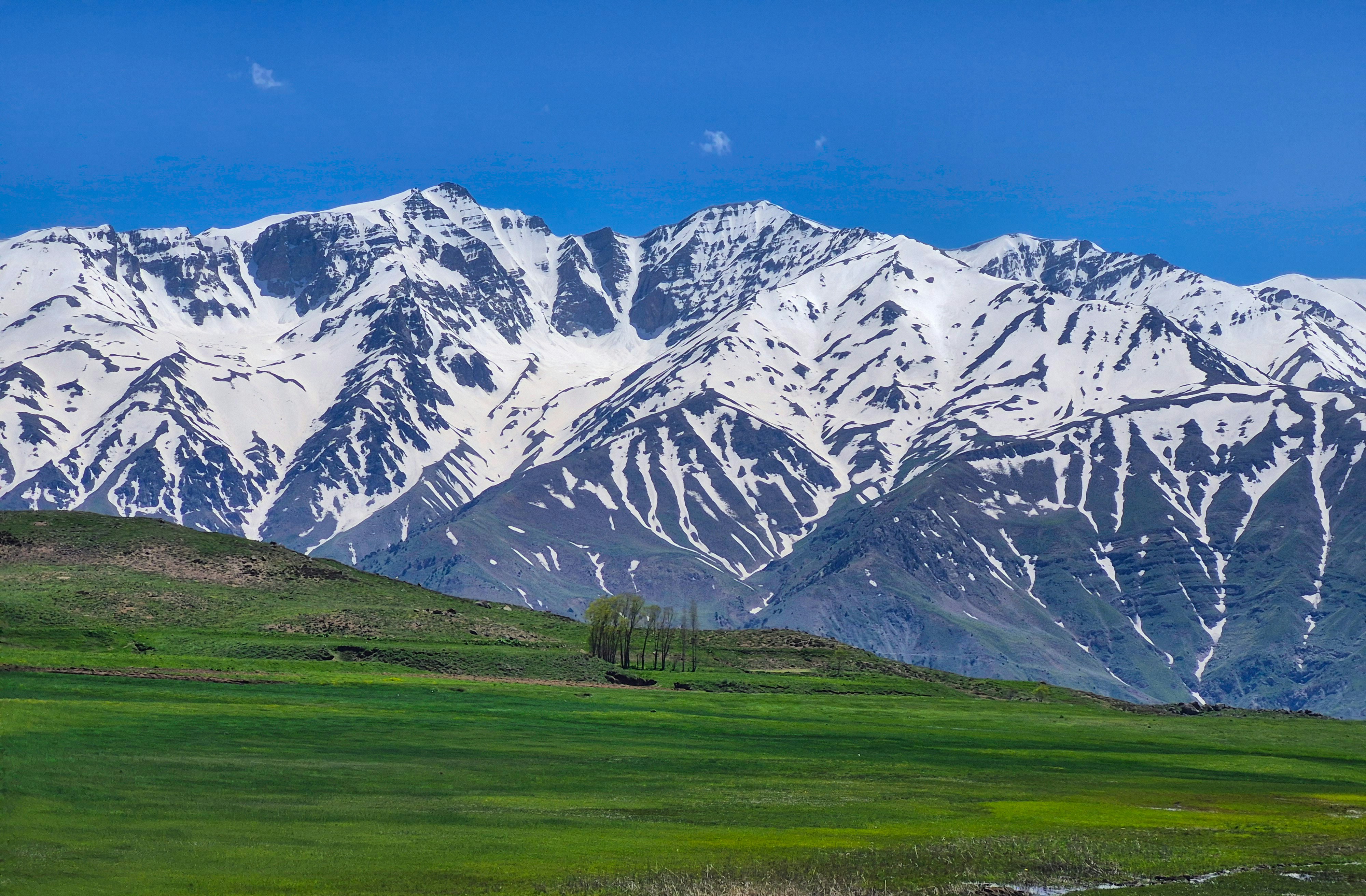 Snow-capped mountains rise above a green hill under blue sky.