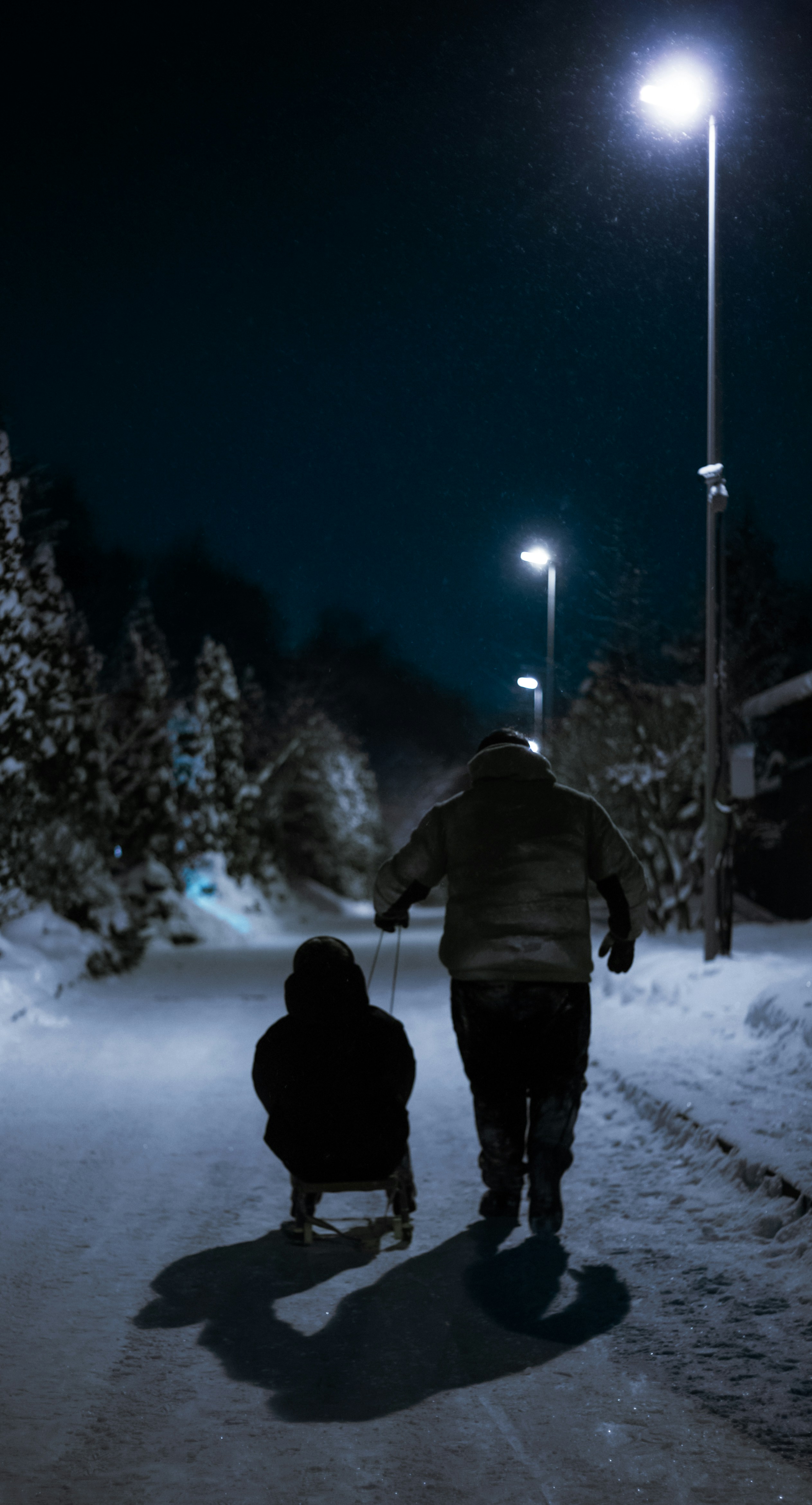 Night Snow Walk: A Lone Silhouette Pulls a Child Through Quiet Street Lights
