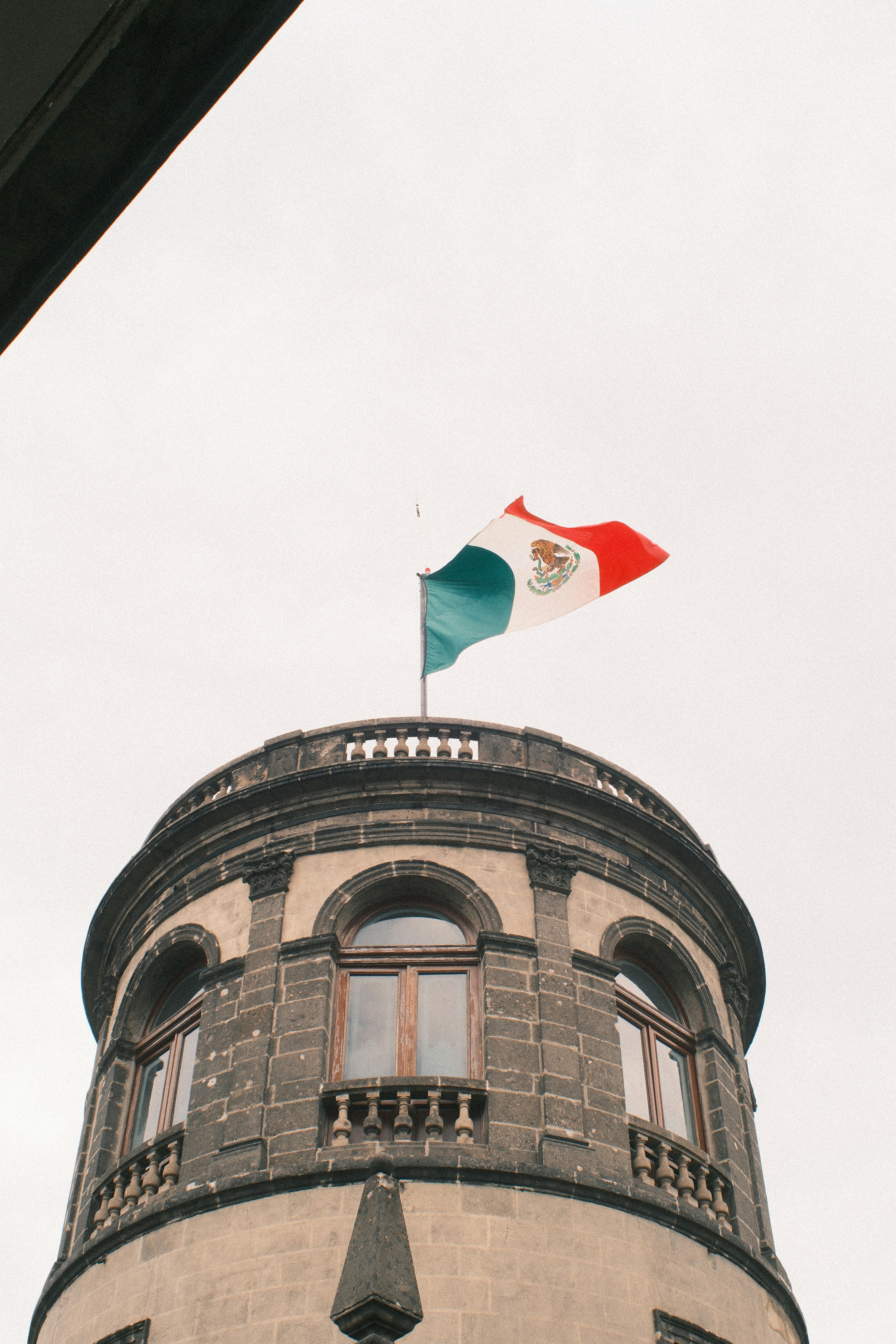 Flag atop circular stone tower: a Mexican flag against a pale sky