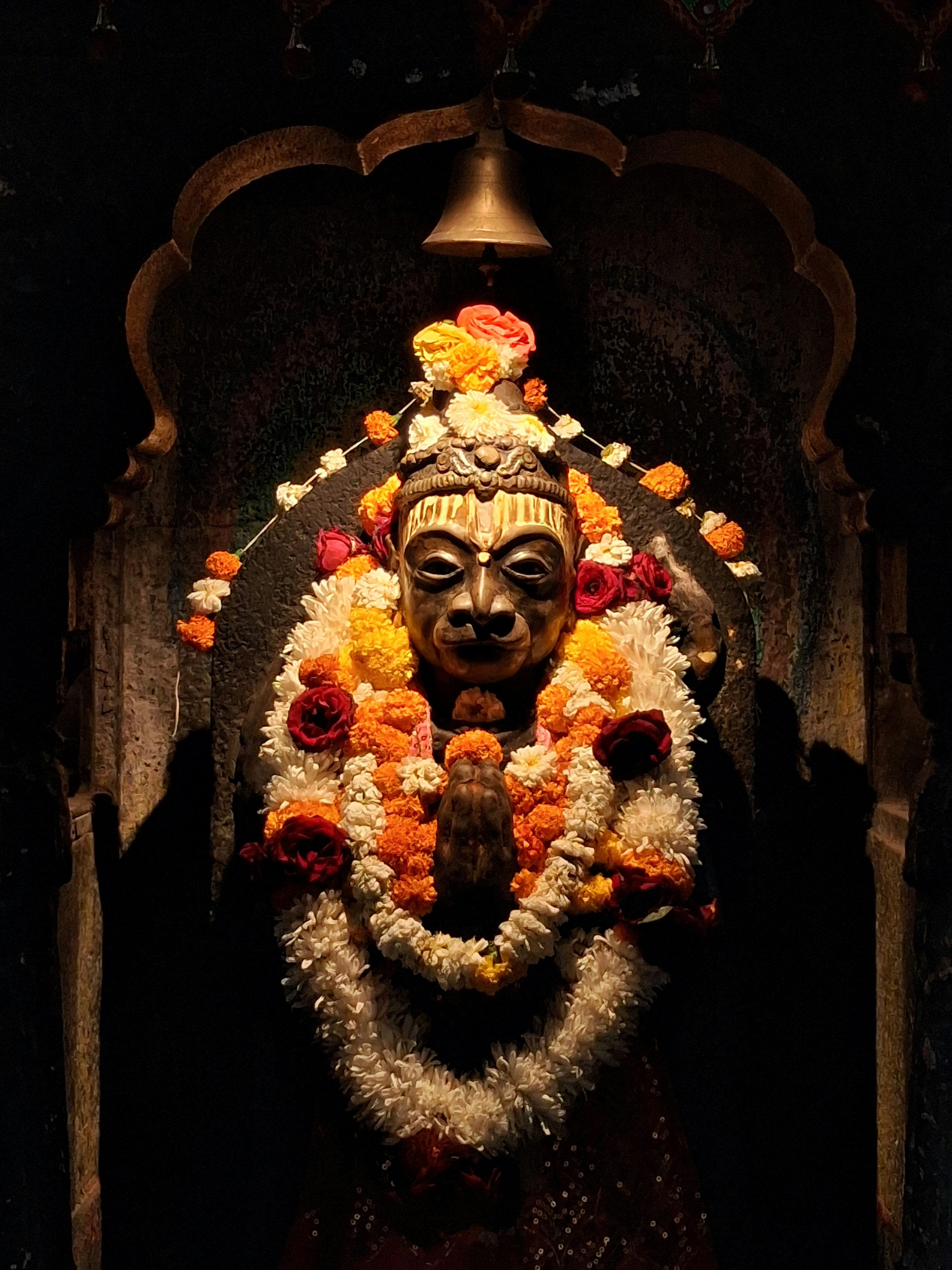 An ancient stone statue of Lord Hanuman, beautifully adorned with fresh flower garlands. Captured in a dramatic low-key lighting style, the golden light highlights the serene and powerful facial features of the deity, while the background remains in deep shadow. A traditional temple bell hangs perfectly above, enhancing the spiritual and mystic essence of Indian heritage. Perfect for themes of spirituality, Hinduism, devotion, and fine art photography.