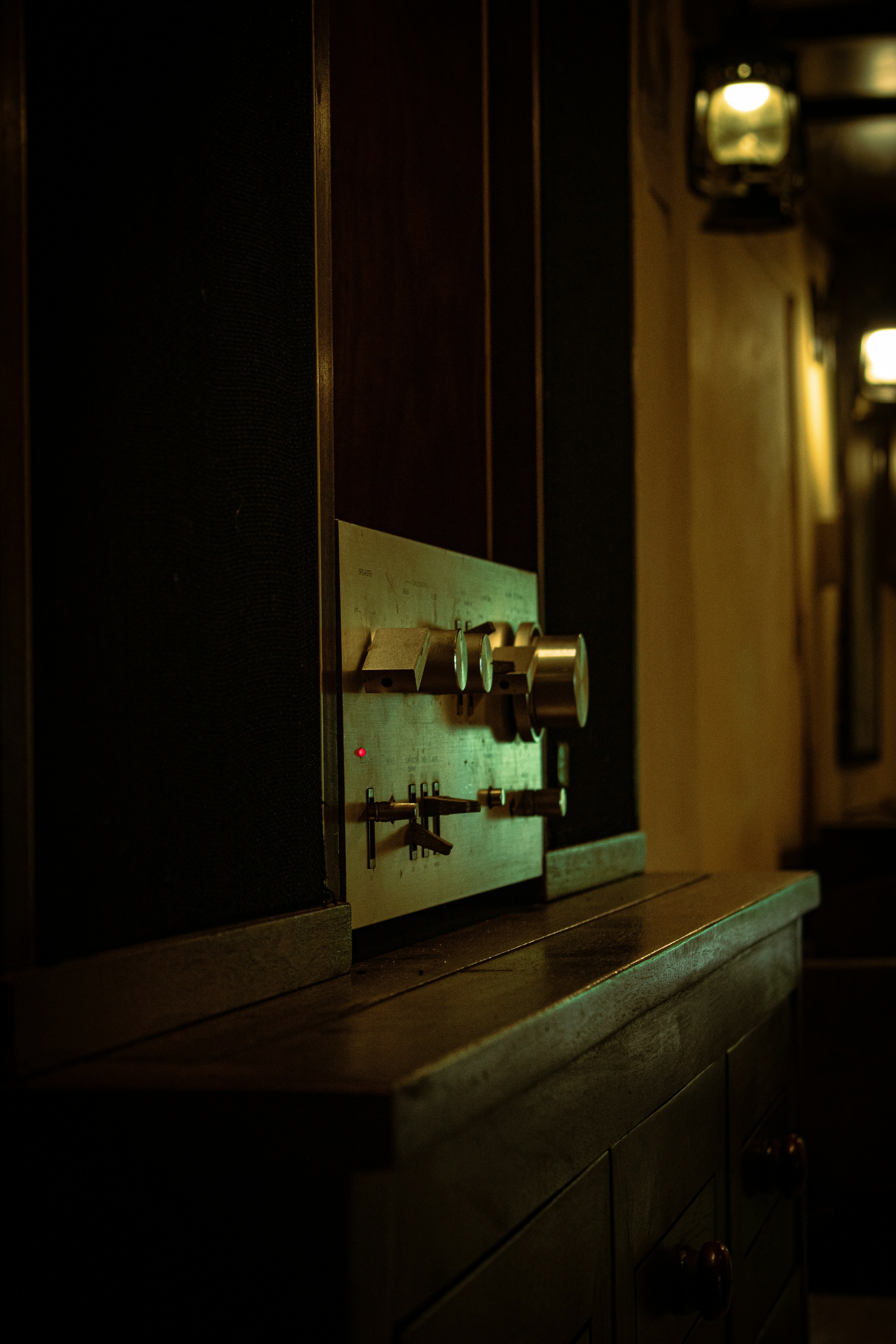 A vintage control panel sits on a dark wooden surface in a dim corridor, illuminated by warm wall lamps and casting soft shadows across the grainy scene.