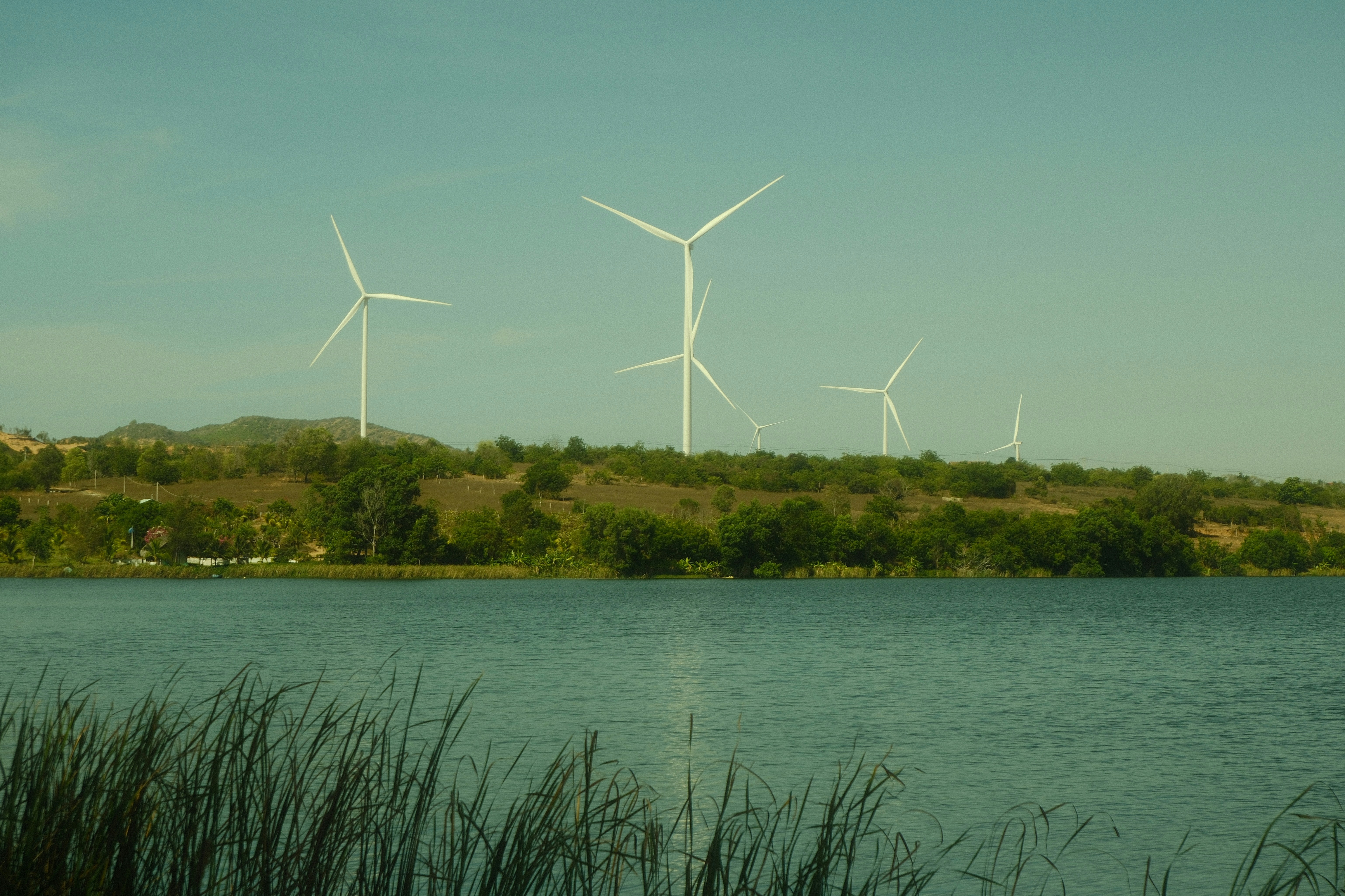 White wind turbines on green hills beside a serene lake.