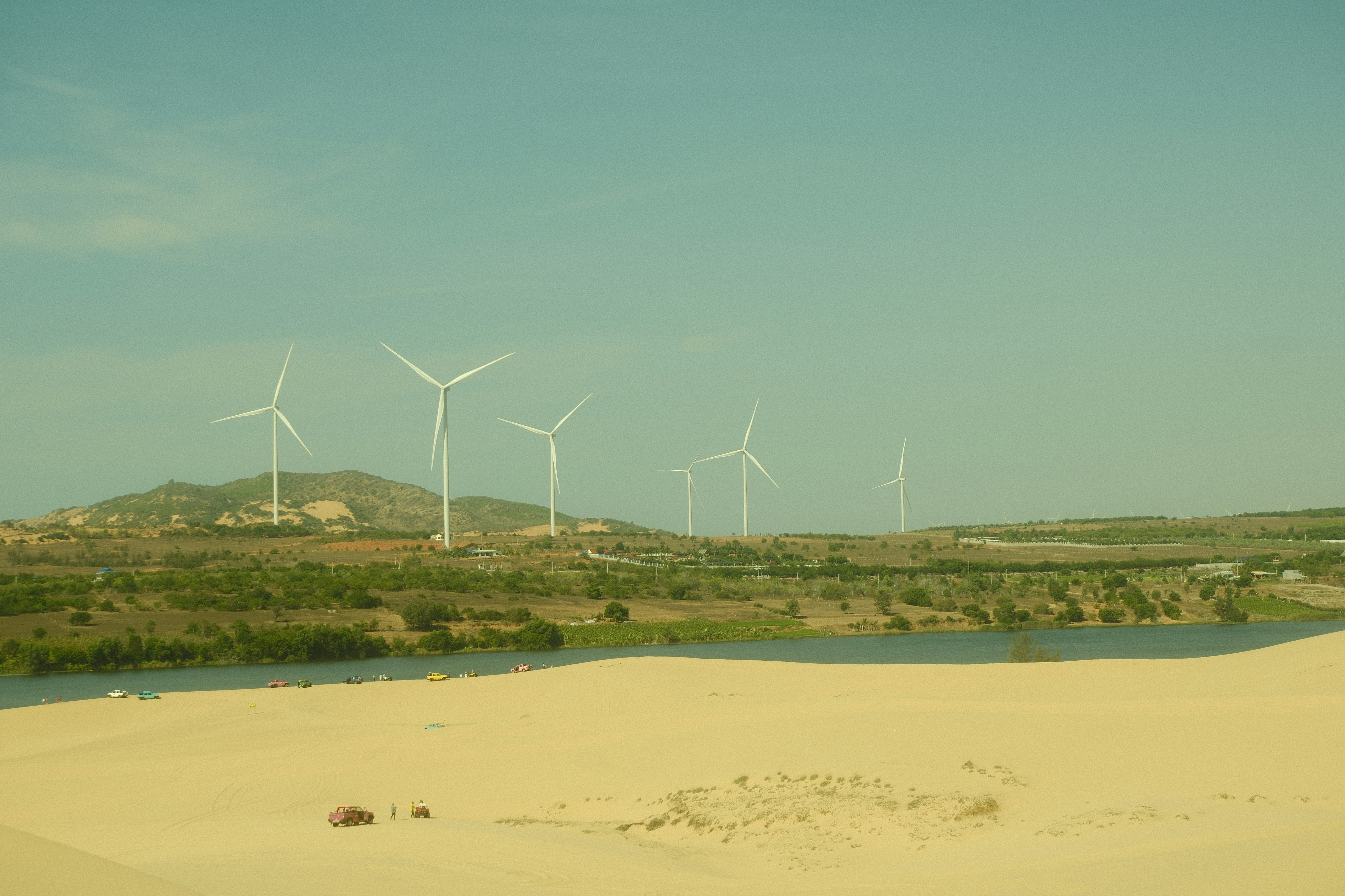 White wind turbines on green hills overlooking a sandy landscape.