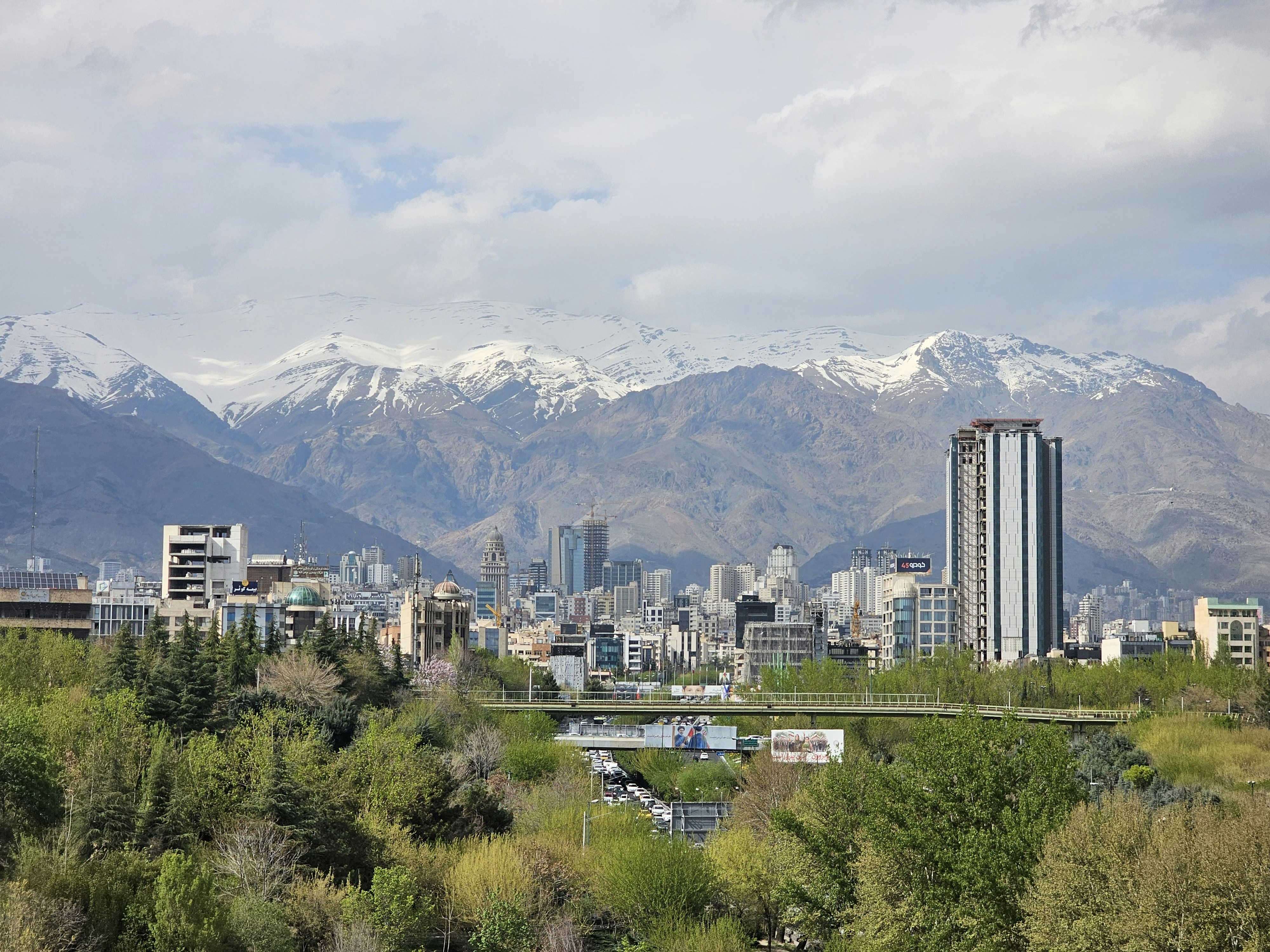 A wide view of Tehran’s skyline rising above fresh spring greenery, backed by the snow-capped Alborz Mountains under a soft, cloudy sky—an urban landscape where city and nature meet.