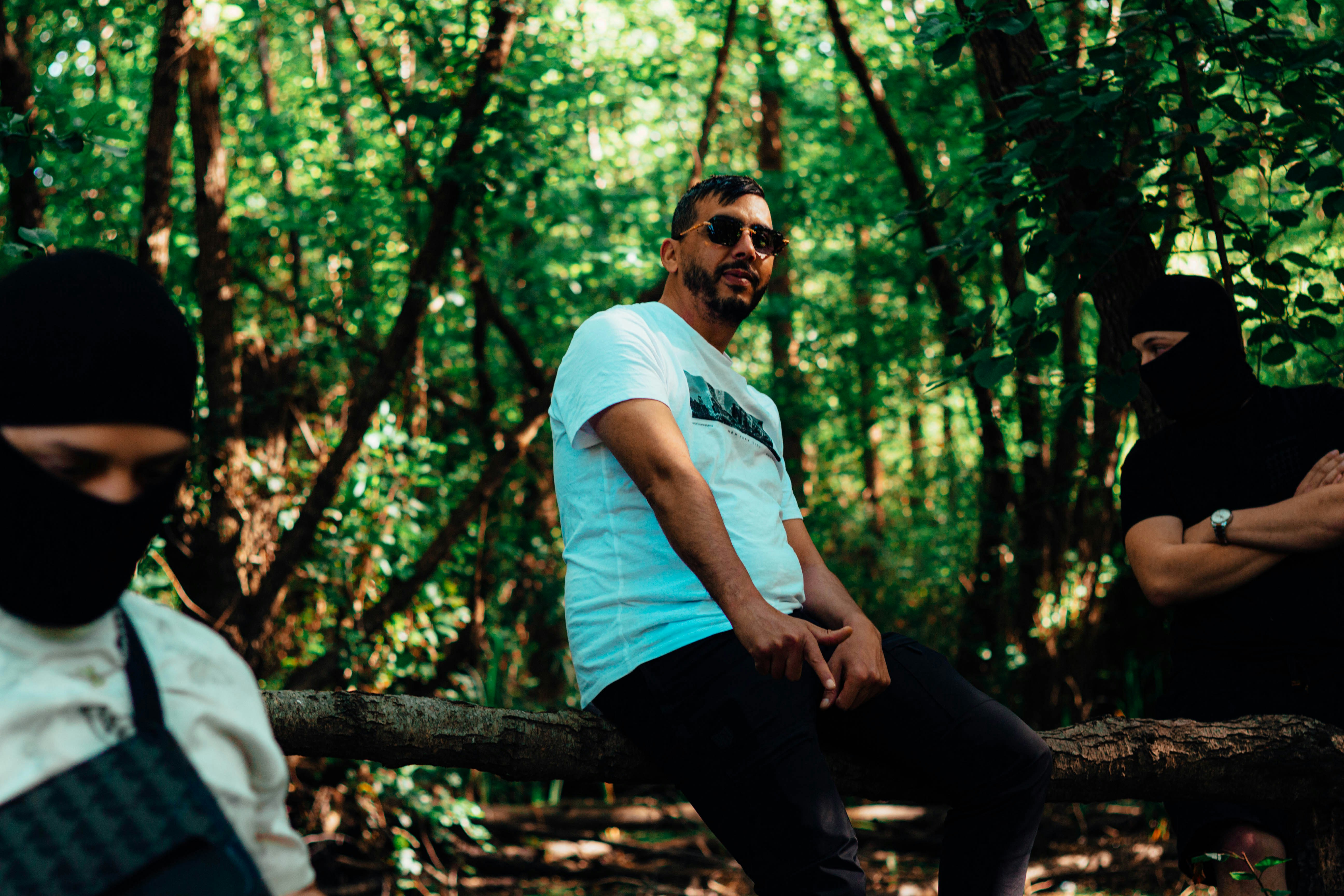 Man on a log amid a sunlit forest, flanked by masked figures