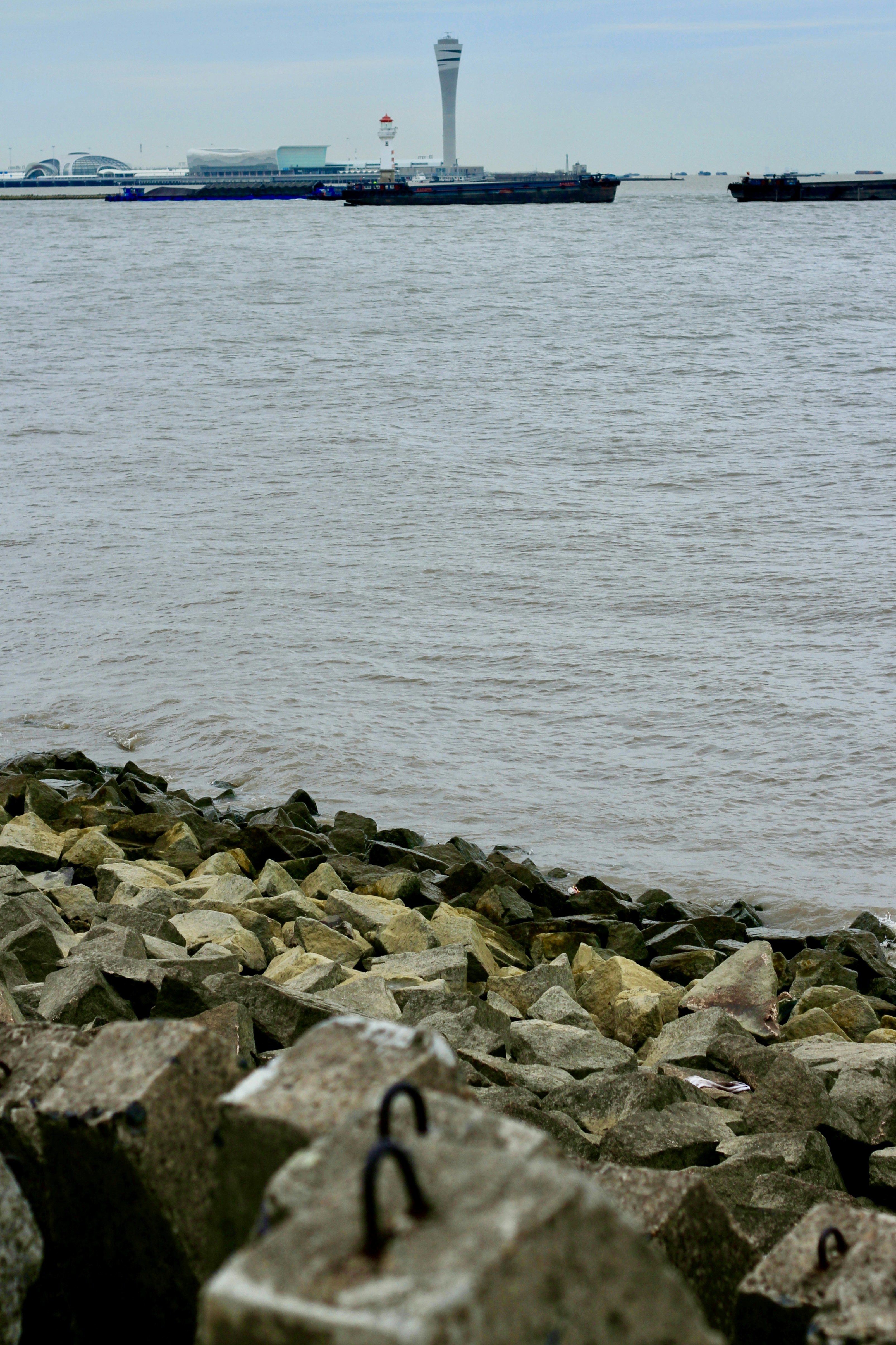 Rocks and breakwater ledge in the foreground with a calm harbor and distant control tower on the horizon.