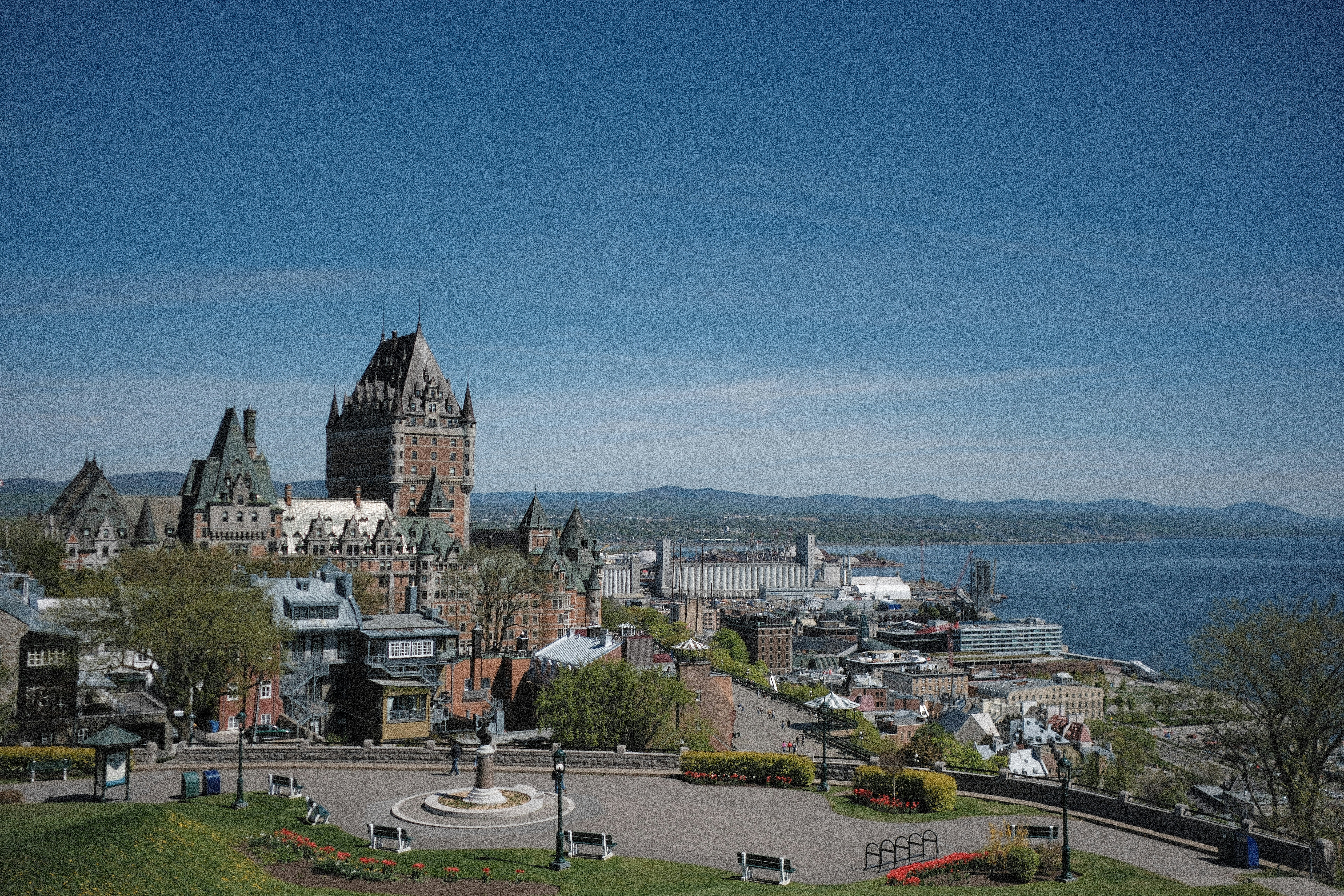 A high-angle landscape view looks out over the historic Upper Town of Quebec City and the sweeping blue waters of the St. Lawrence River. The iconic Fairmont Le Château Frontenac stands proudly on the left, resembling a grand castle with its complex brick walls and steep green roofs. Below the fortress, a public promenade features green lawns, small flower patches, and neat white benches for visitors. Far across the blue river, a detailed city coastline stretches under an open sky. This photo was taken by Fujifilm xt5 with 23mm and no edit just fuji colors.