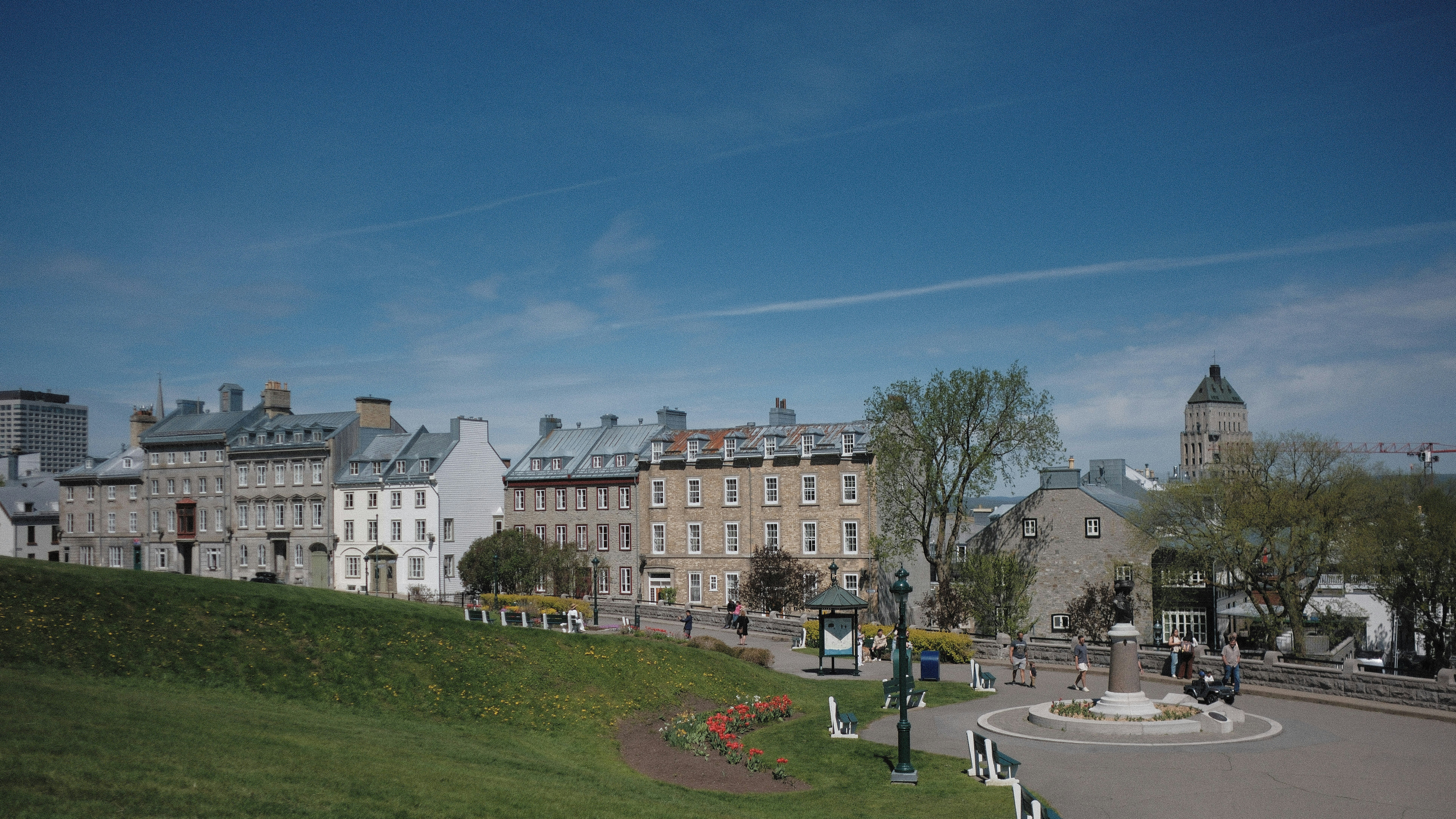 A beautiful public square in Quebec City sits alongside a lush green hill under a vast blue sky. Classic stone houses with dark metal roofs line the background, showcasing the beautiful art of historic neighborhood design. In the foreground, neat park benches and a small monument fountain are surrounded by a curving garden bed of red tulips. This photo was taken by Fujifilm xt5 with 23mm and no edit just fuji color, perfectly preserving the authentic, bright midday textures and sunny outdoor mood.