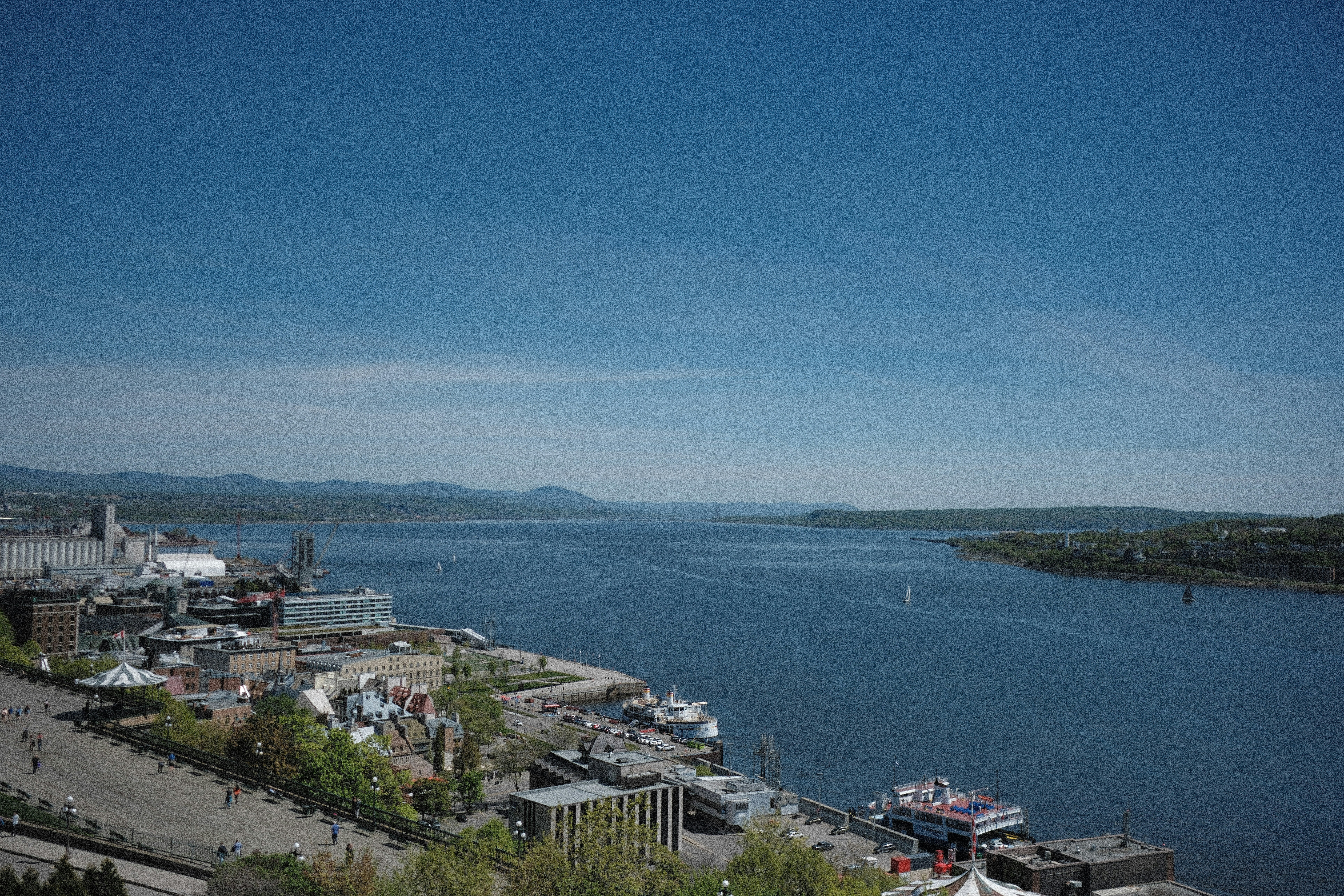 A vast, high-angle panoramic photograph displays the expansive blue St. Lawrence River stretching out to the horizon. In the lower left, the charming rooftops of Old Quebec and a long pedestrian promenade frame the water. Several boats, including a large passenger ferry and small distant sailboats, glide across the surface under a soft sky. This photo was taken by Fujifilm xt5 with 23mm and no edit just fuji color, showing off the beautiful, unedited scale of the river landscape and the peaceful art of coastal travel.