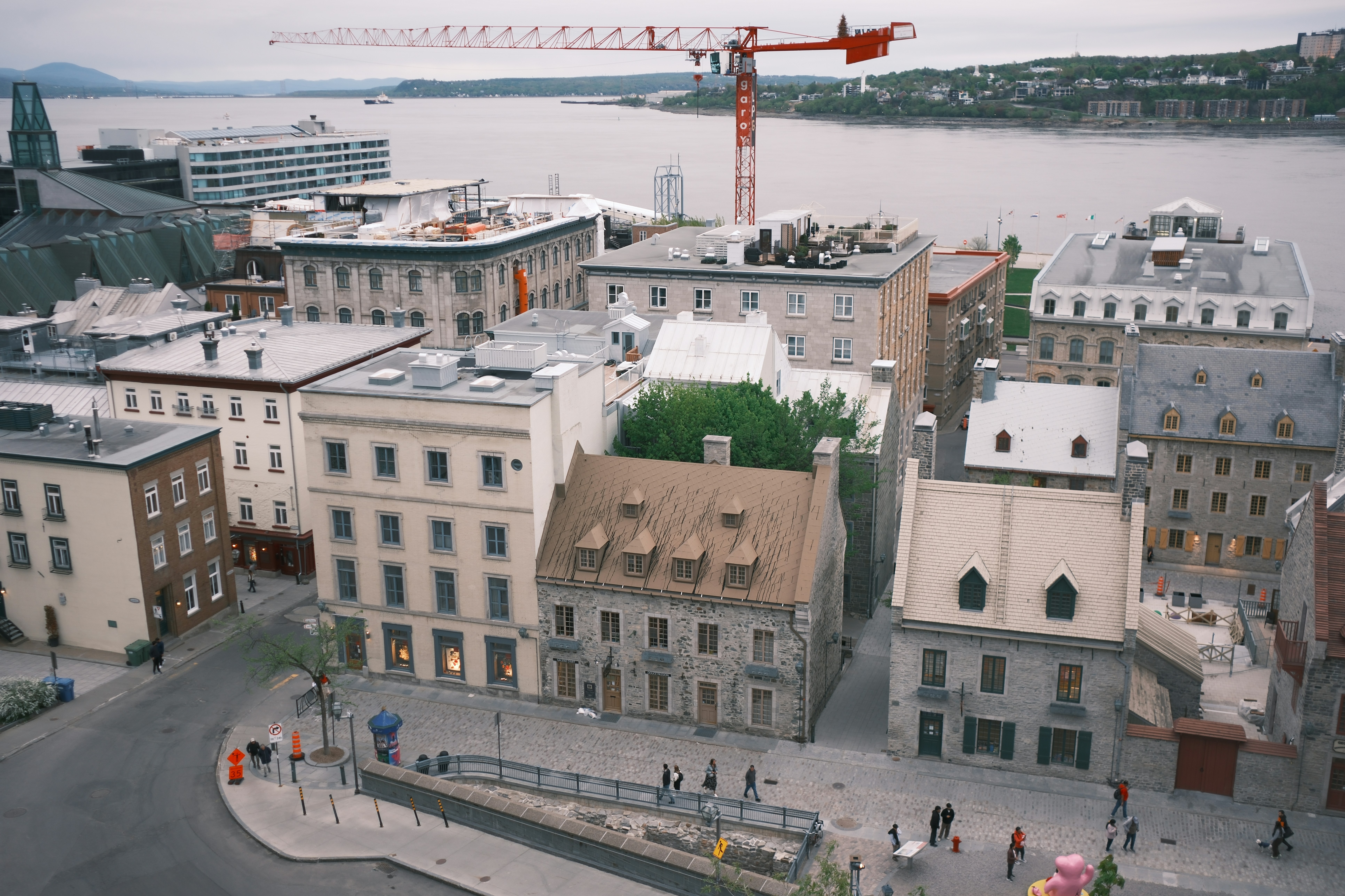 A high-angle landscape photograph looks down on the charming rooftops and historic cobblestone plazas of Old Quebec. Classic stone houses with pitched roofs stand tightly packed next to a curved pedestrian walkway where several small groups of people are walking. In the background, a large red construction crane stands tall over the city blocks near the wide, misty waters of the St. Lawrence River. This photo was taken by Fujifilm xt5 with 23mm and no edit just fuji color, perfectly preserving the authentic, unedited tones, rooftop layouts, and grand coastal scenery.