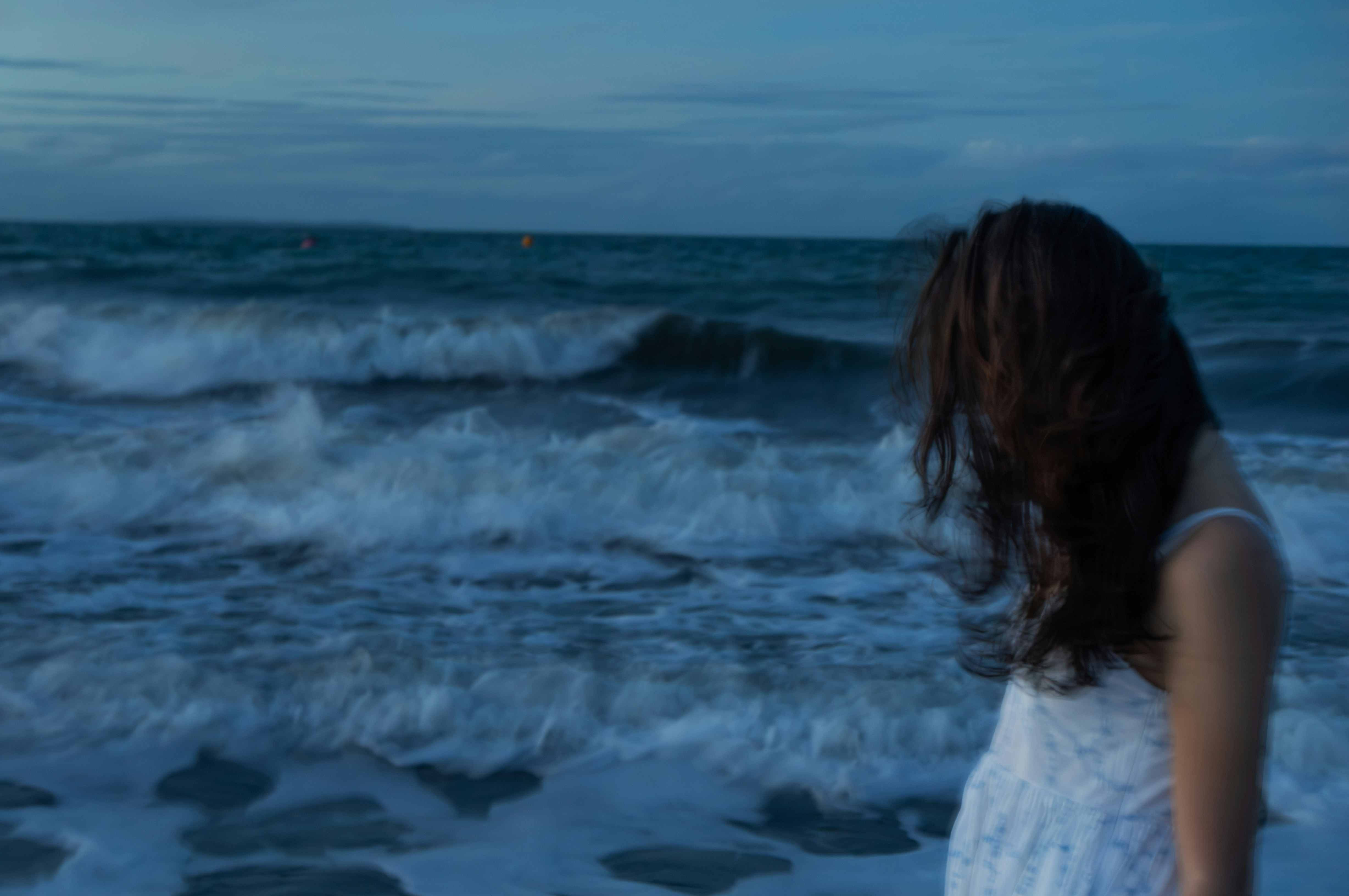A woman with dark hair stands by the wavy ocean.